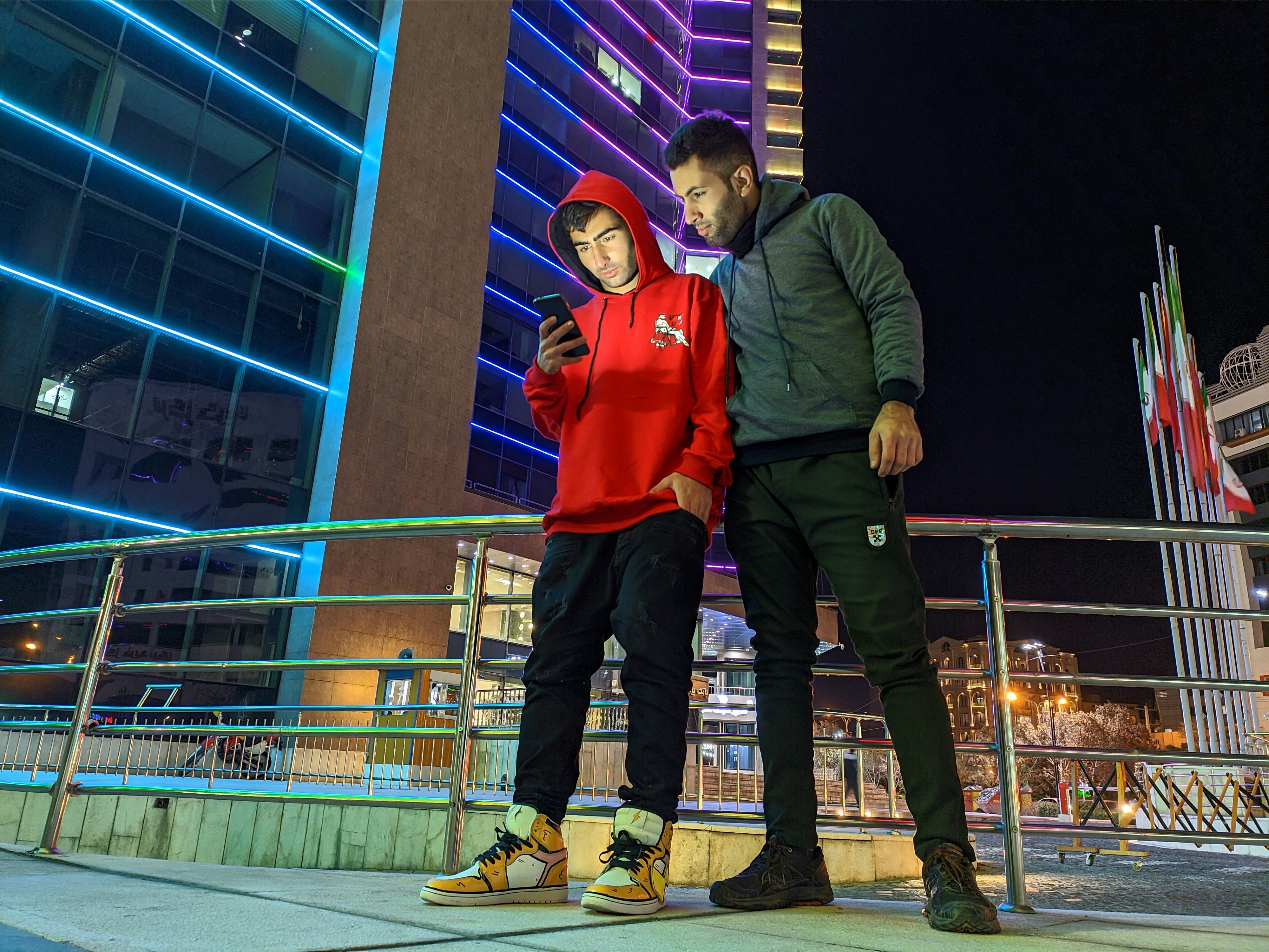 Two friends walk along a railing at night beneath neon-lit skyscrapers, the one in a red hoodie checking a phone.