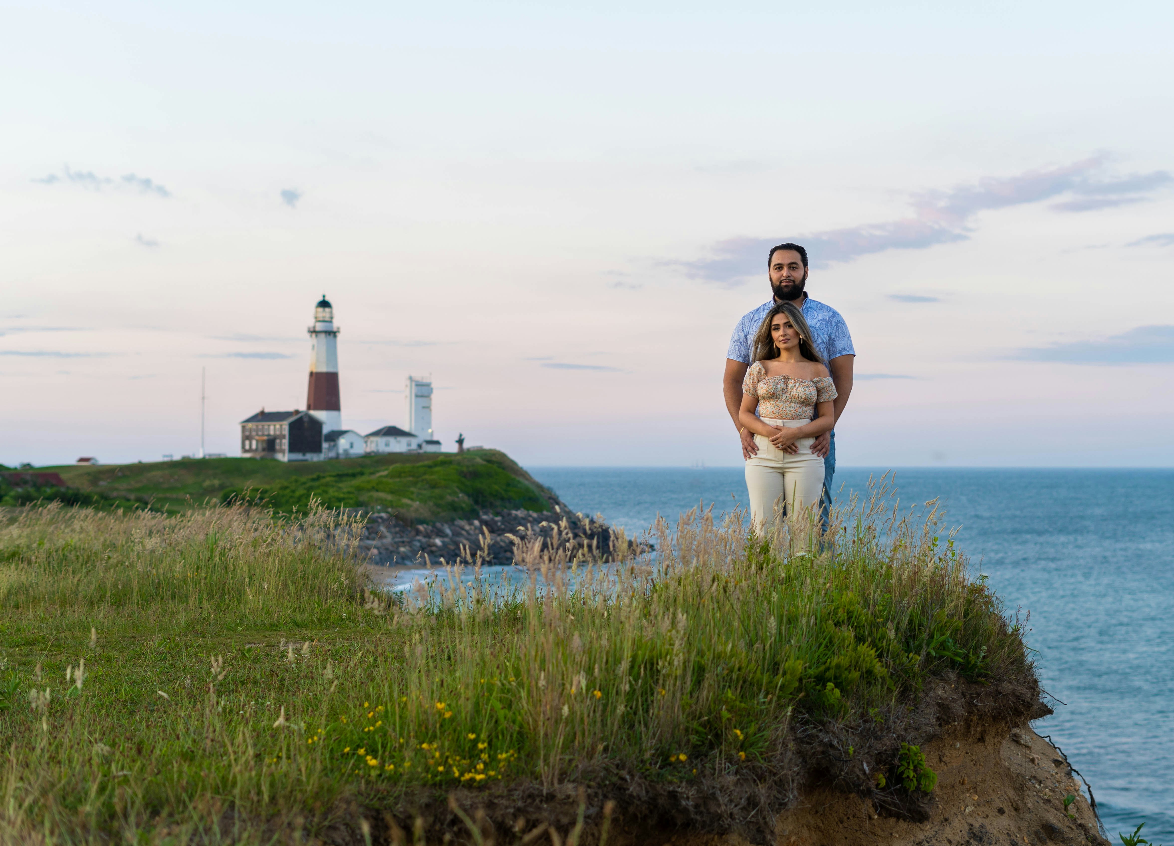 Couple standing together on a grassy cliff, overlooking a lighthouse by the ocean at twilight.