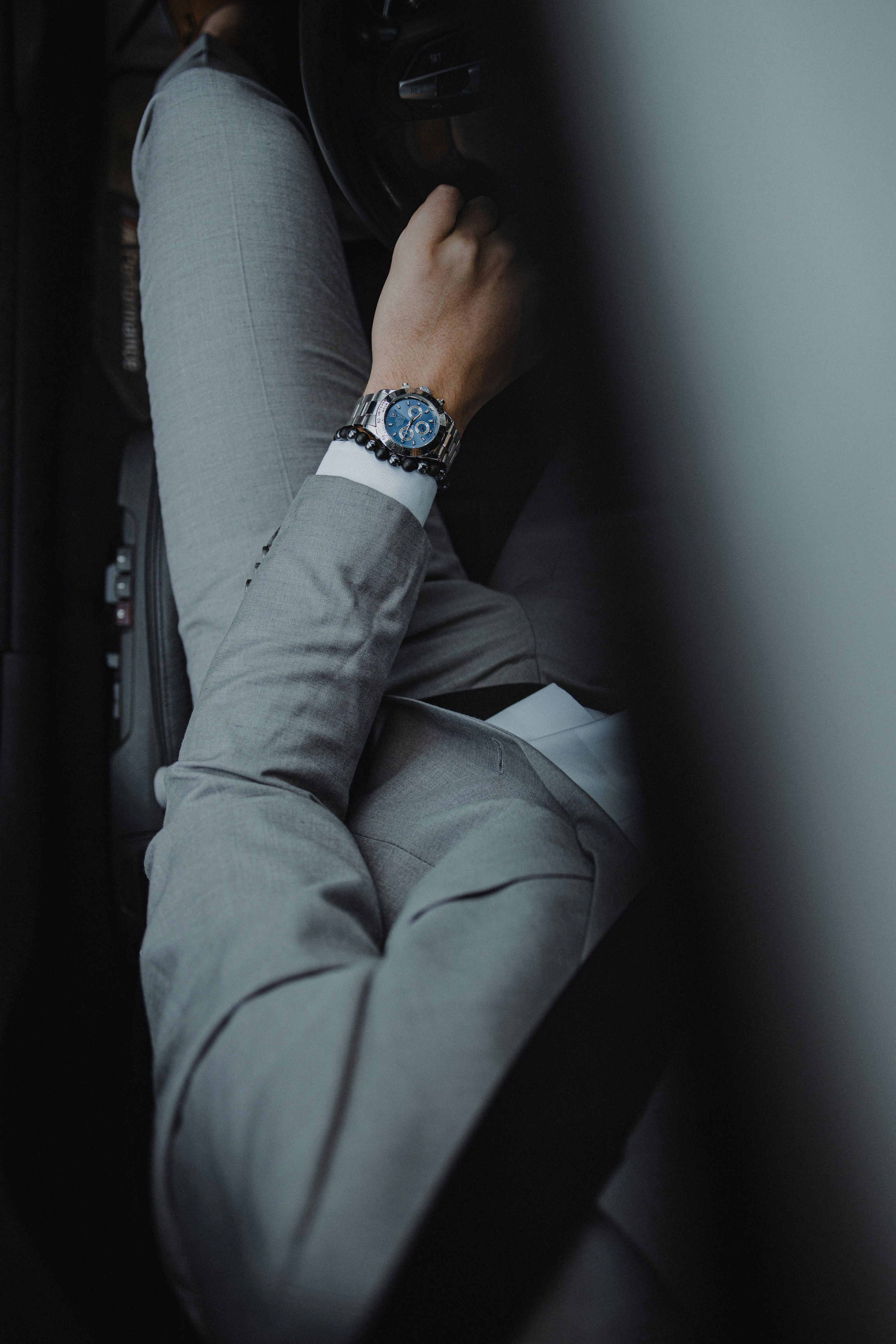 A well-dressed individual in a gray suit relaxes in a car, showcasing a sophisticated watch. The scene captures a moment of luxury and style.