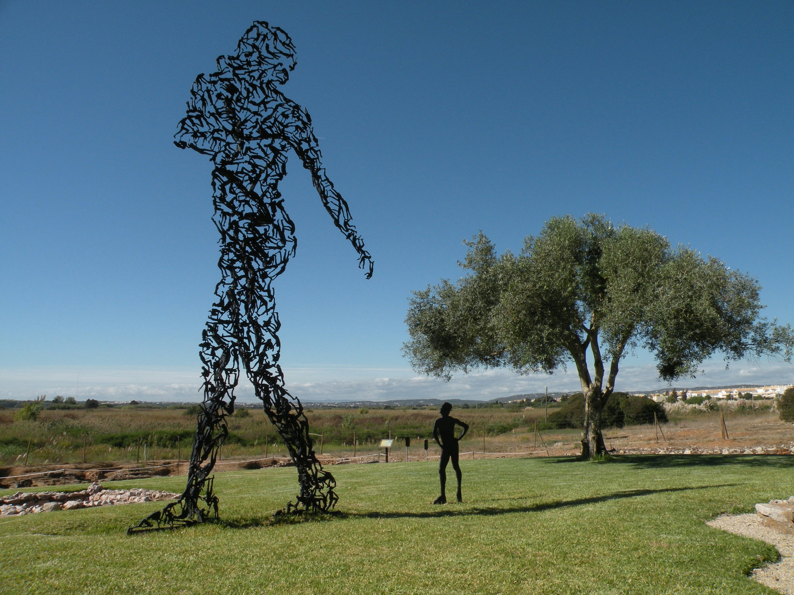 Large wireframe sculpture of a walking figure contrasted against a smaller silhouette, set in a grassy landscape with a lone tree.