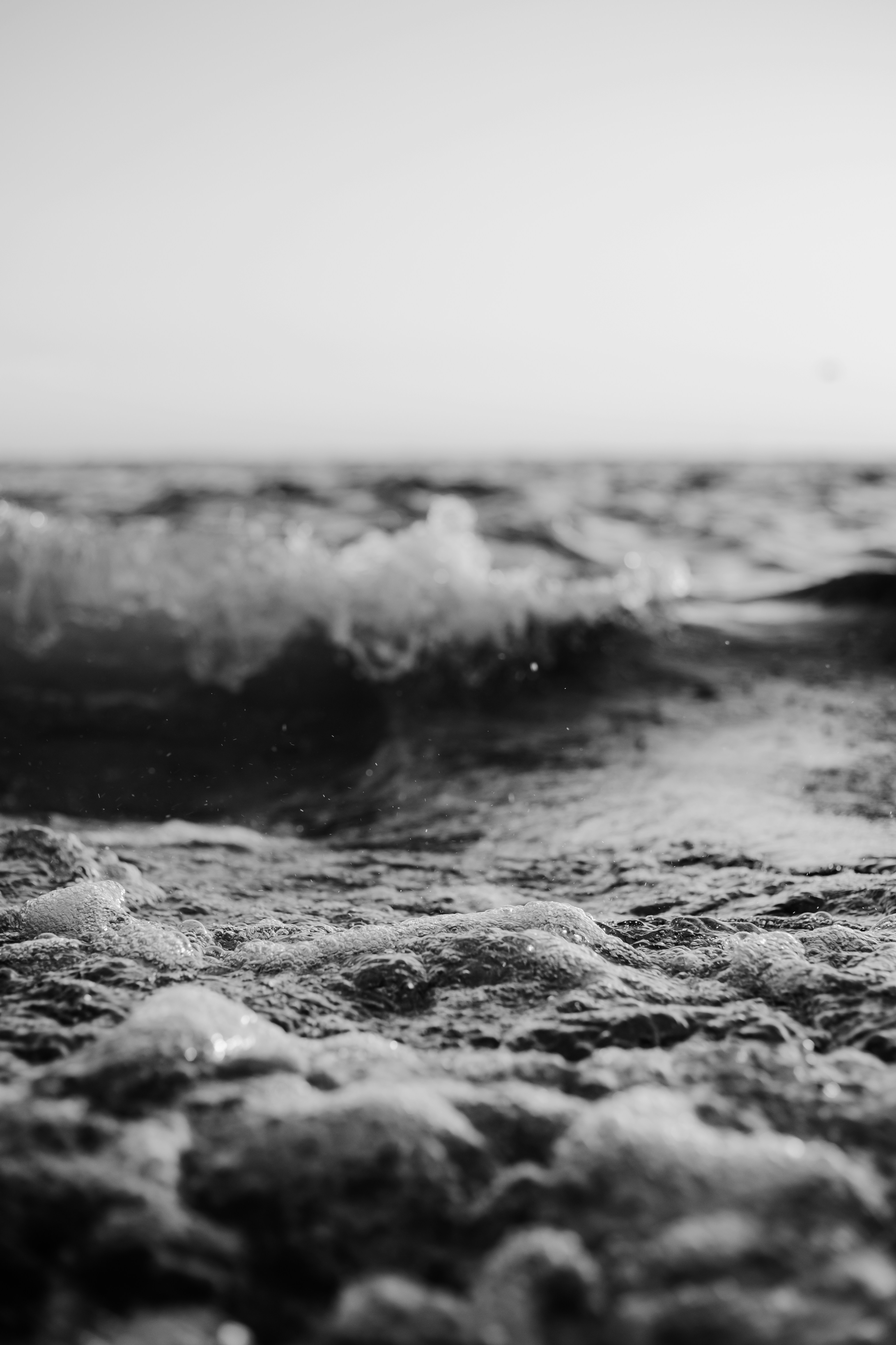 Close-up view of ocean waves crashing against the shore, captured in black and white. The image highlights the texture of the water and foam.