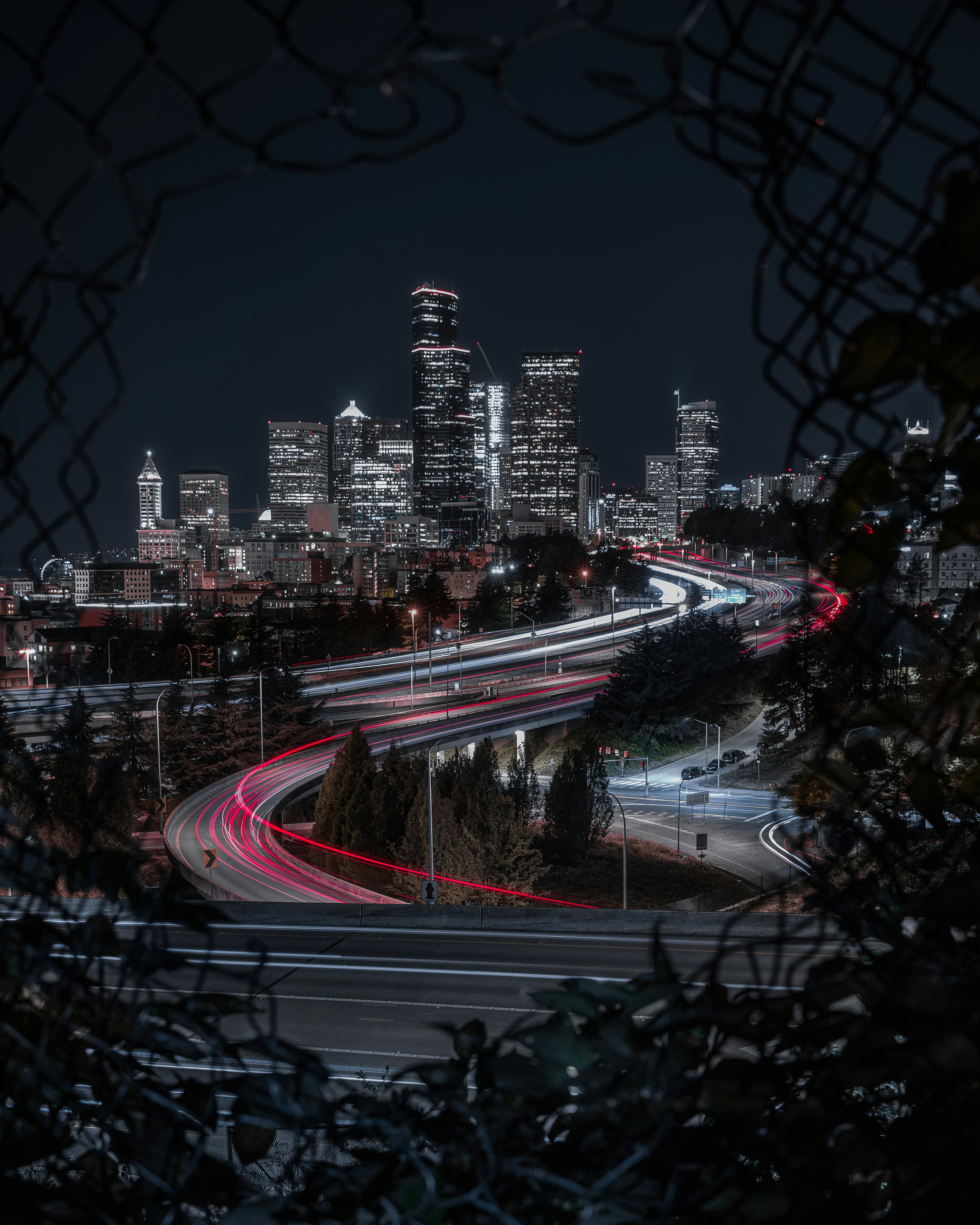 Neon Arc Over the Freeway: City Skyline Framed by a Chain-Link Veil