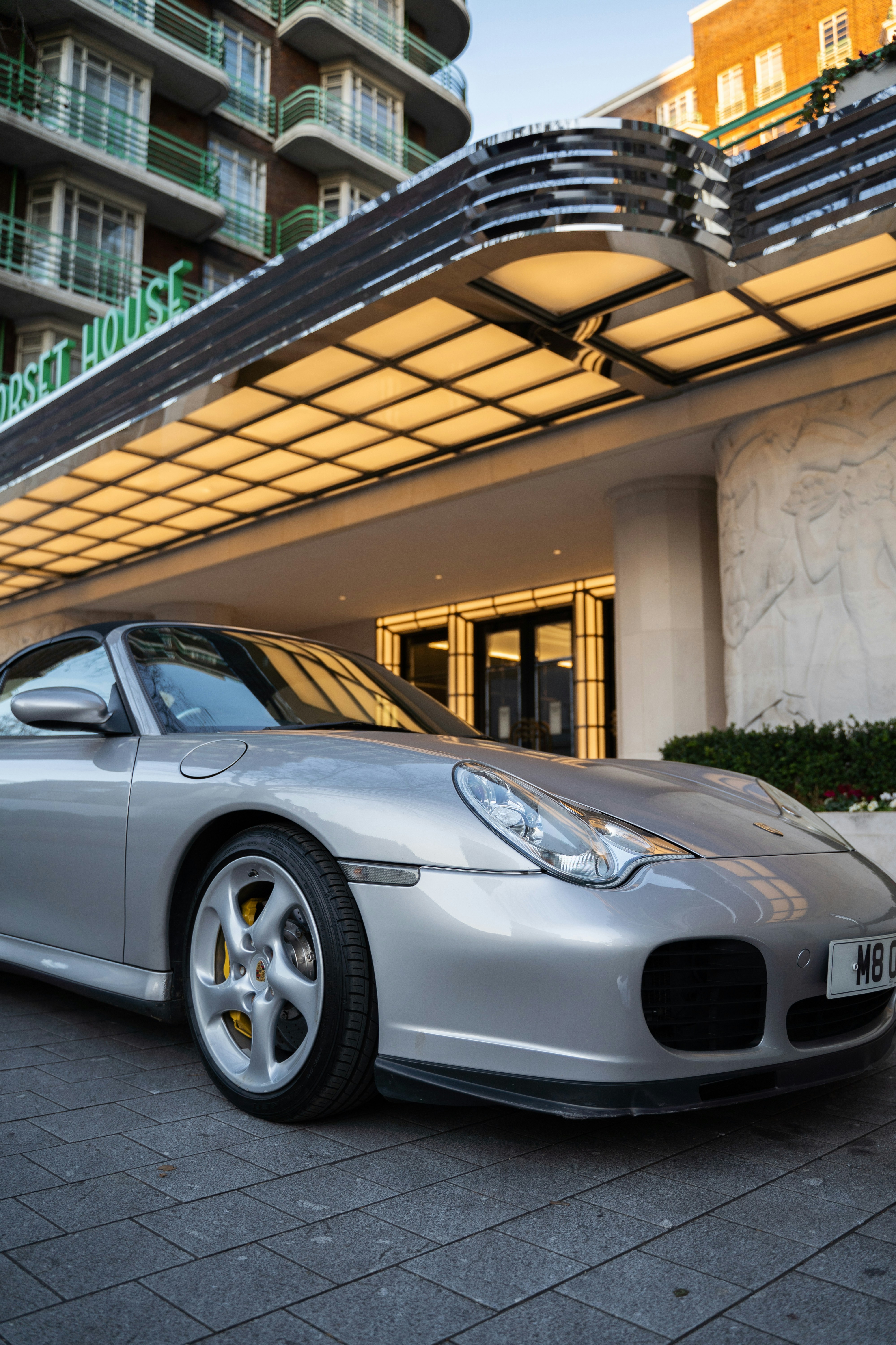 Silver Porsche 911 parked in front of a modern building with art deco features, showcasing its sleek design against a stylish backdrop.
