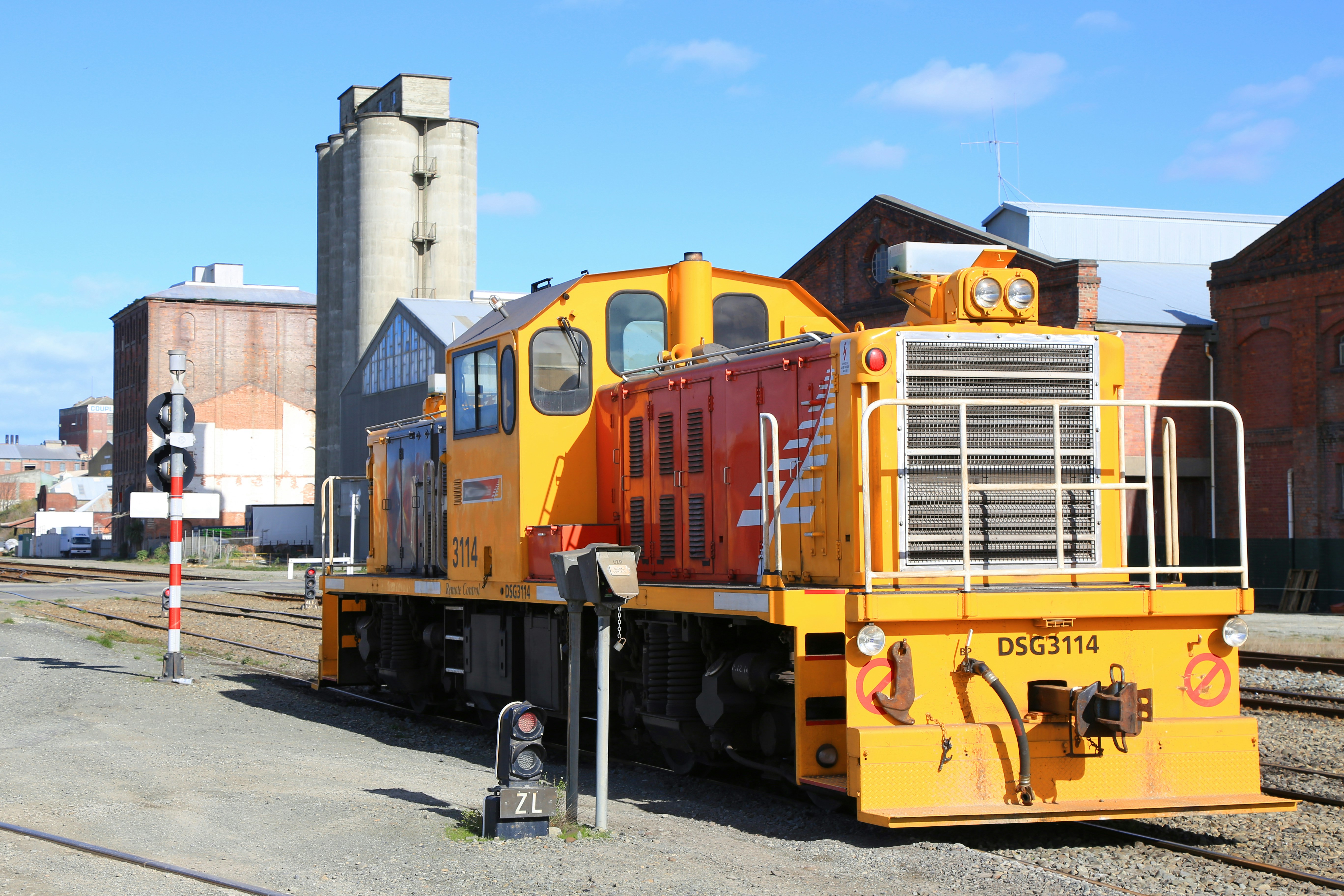 A vibrant yellow locomotive stands idle beside railway tracks, framed by industrial buildings under a clear blue sky.