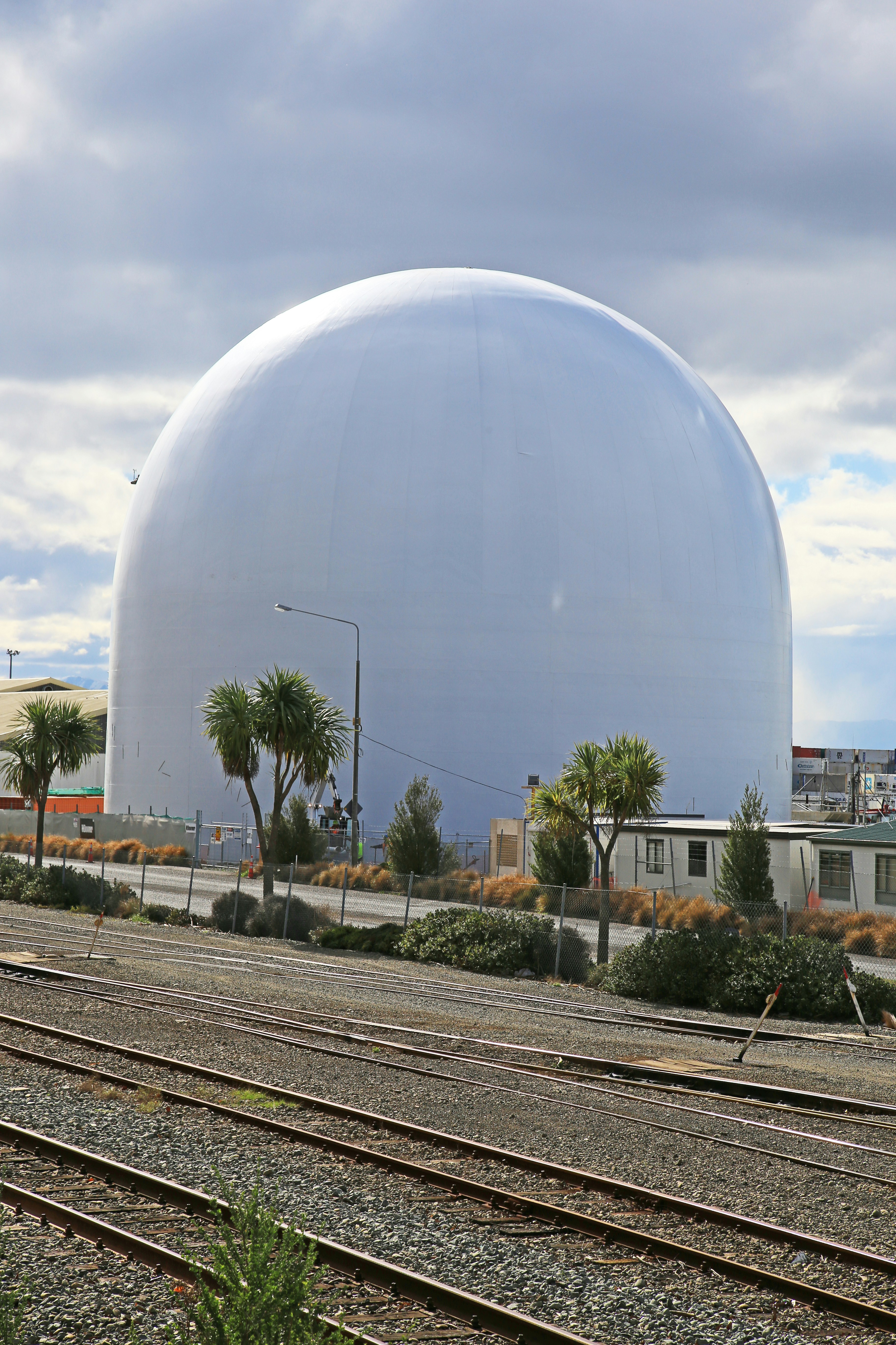 Large, dome-shaped structure surrounded by greenery and railway tracks under a cloudy sky.