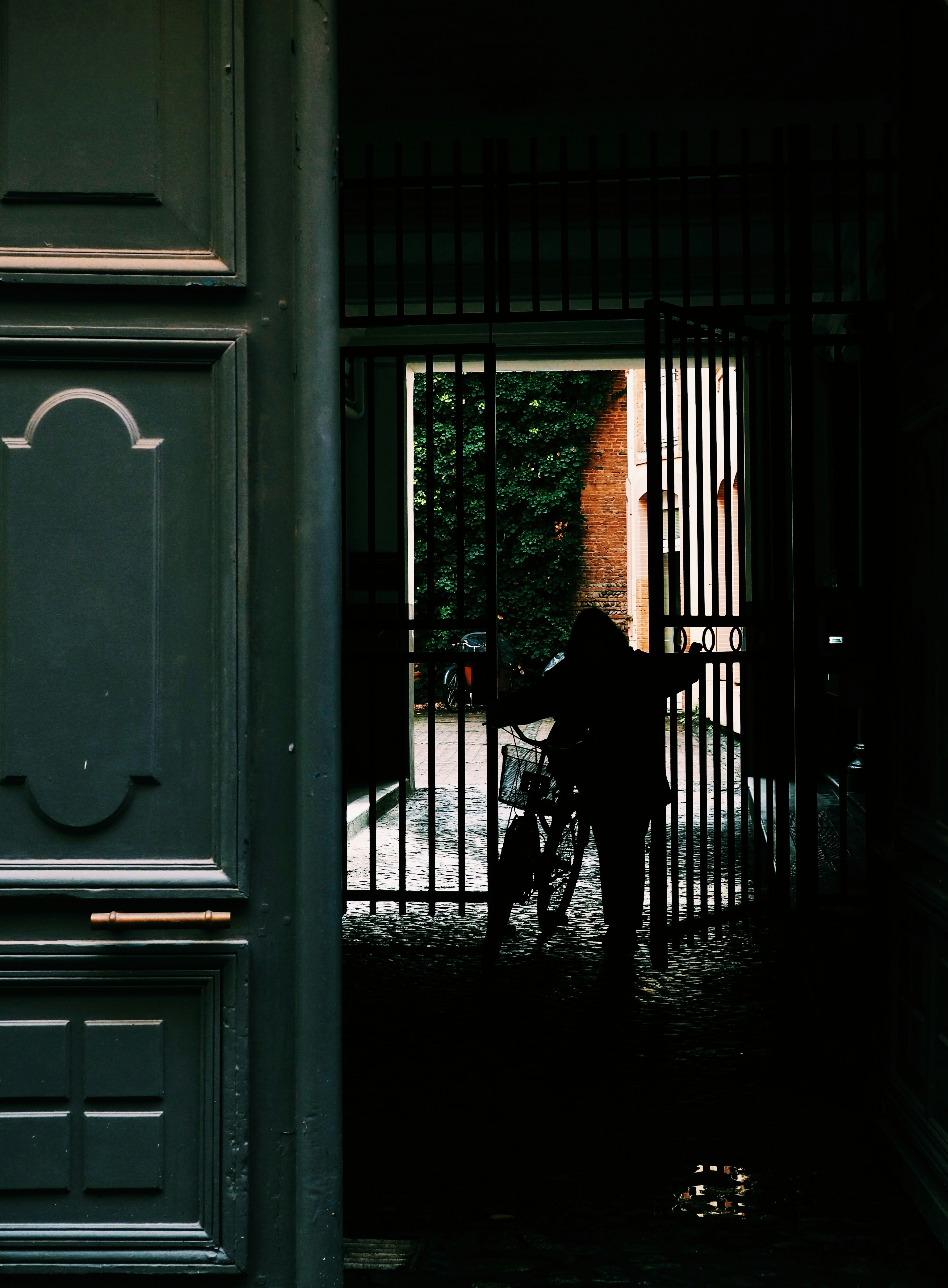 Silhouette of a person pushing open a gate while holding a bicycle, framed by a dark doorway and illuminated courtyard beyond.