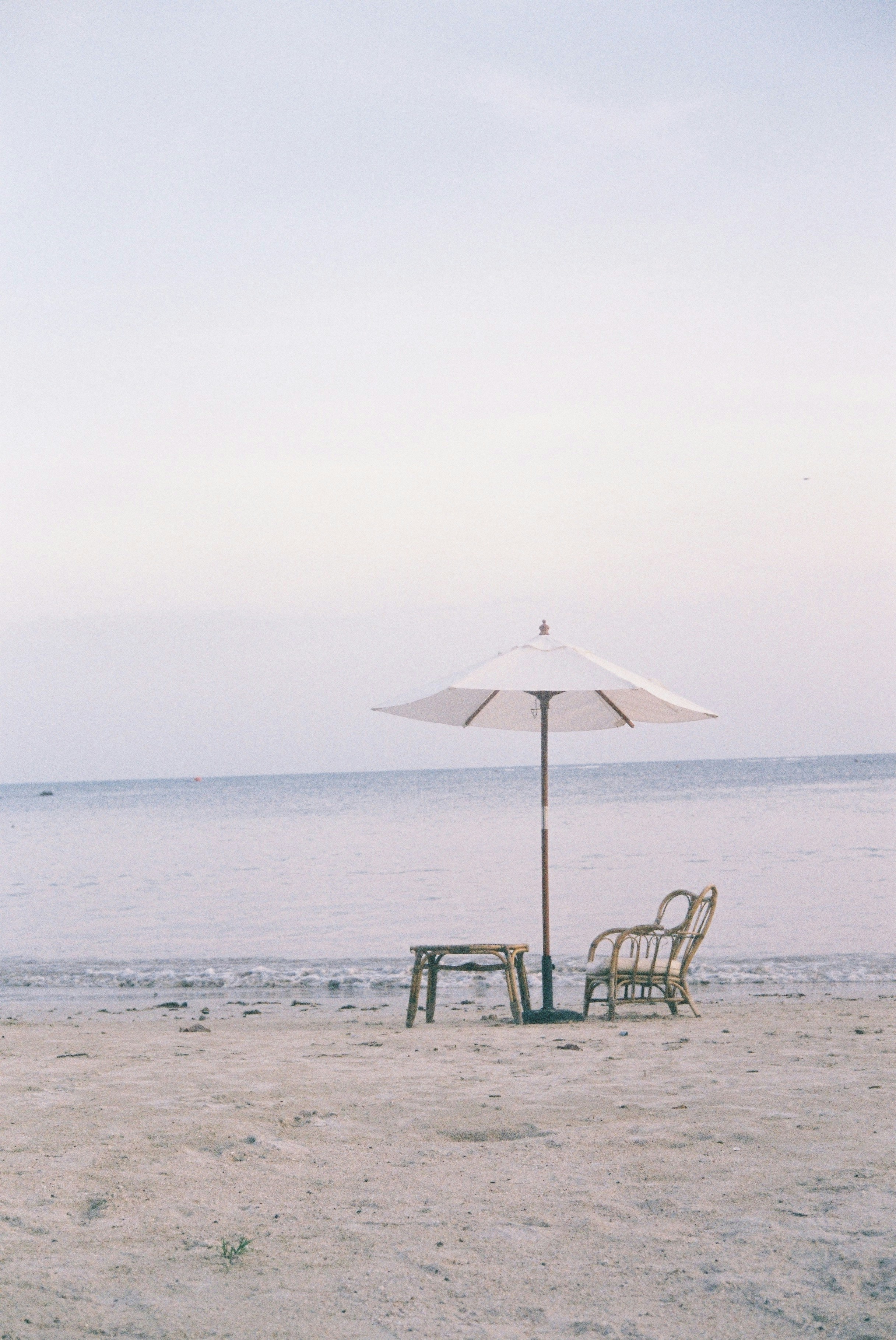 Wooden chair and table under a white umbrella on a sandy beach at the water's edge.