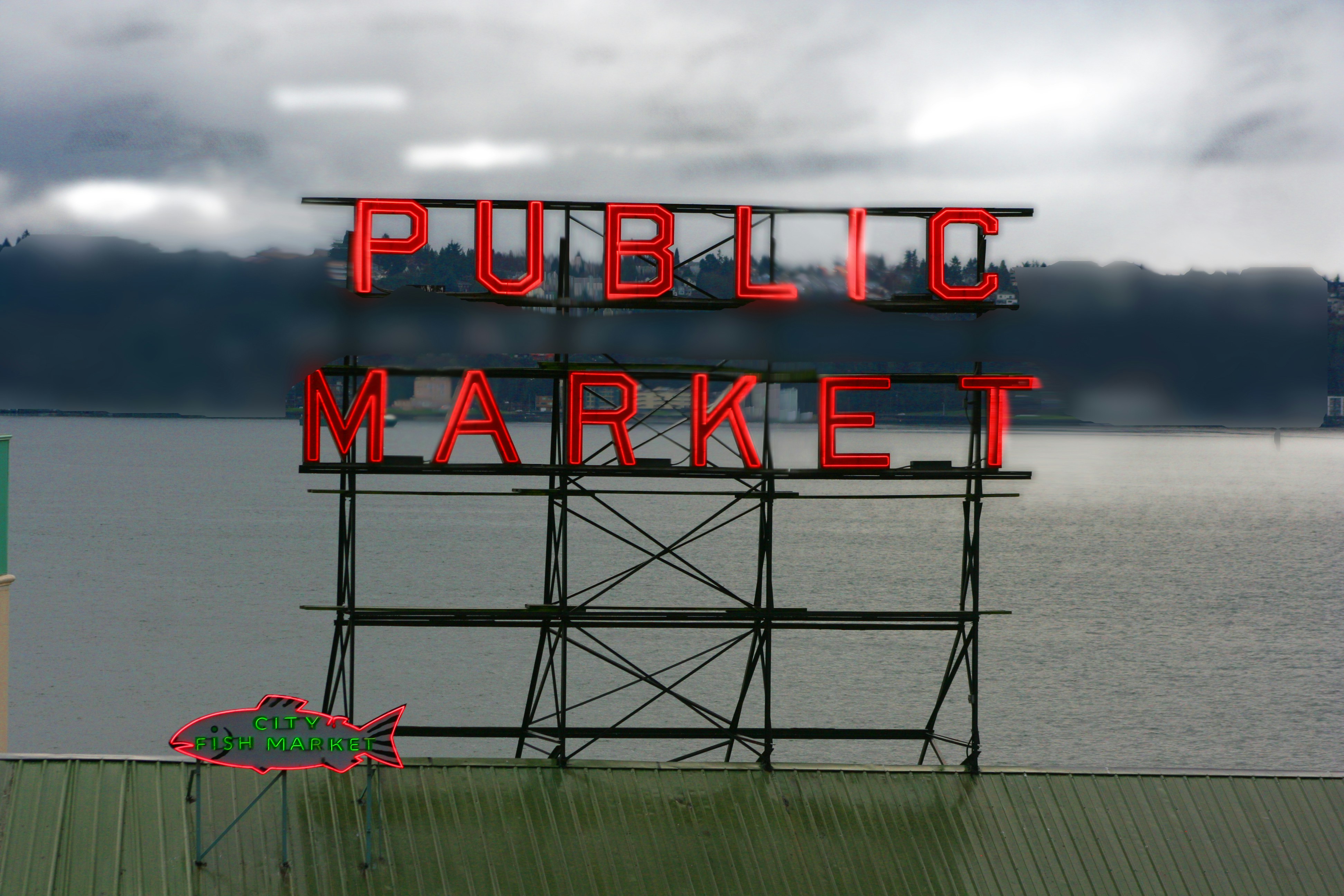 Illuminated 'PUBLIC MARKET' sign atop a structure overlooking water, with a stylized fish logo on the side.