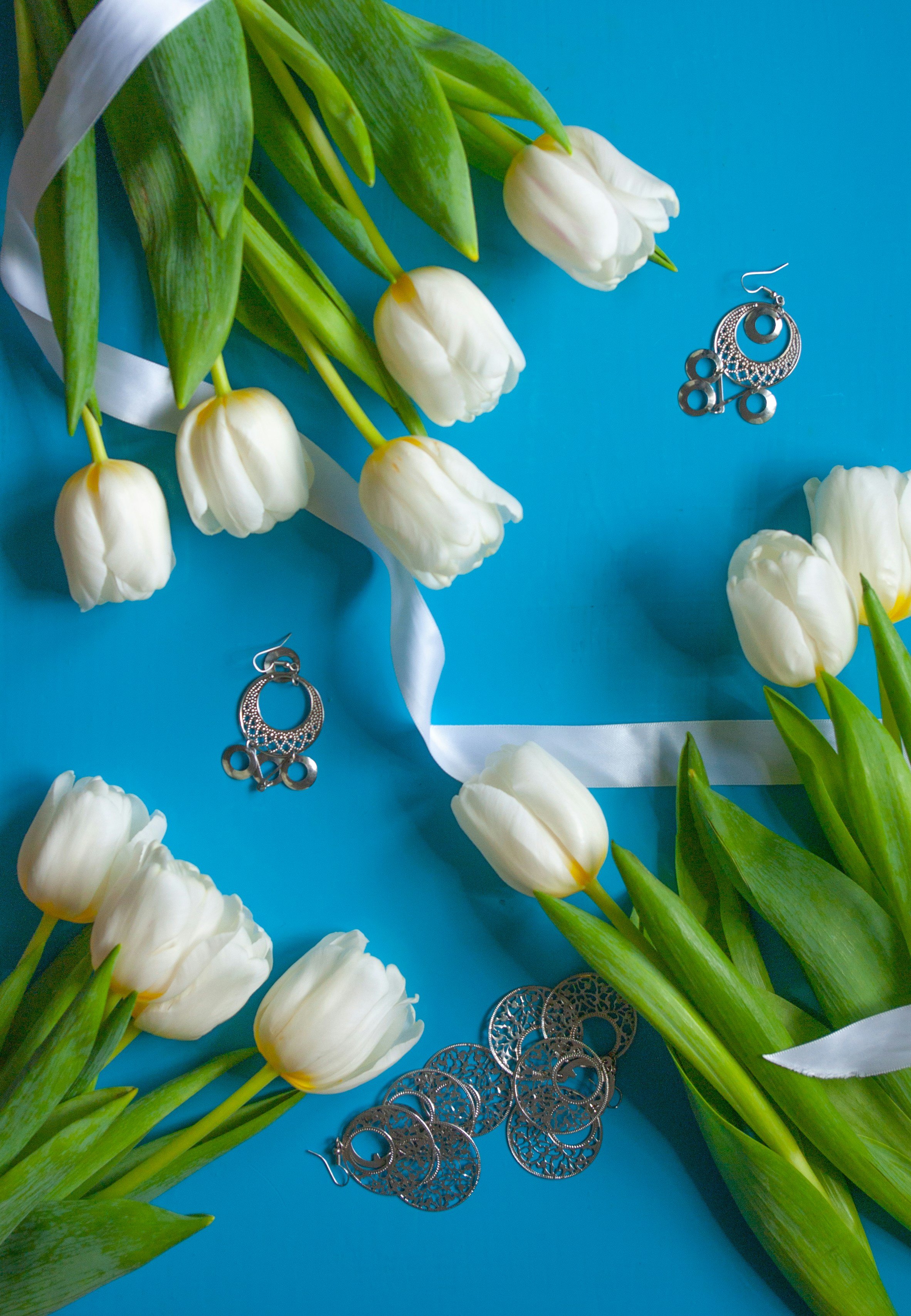 Still life photograph featuring white tulips, silver earrings, and a white ribbon arranged on a vivid blue backdrop.