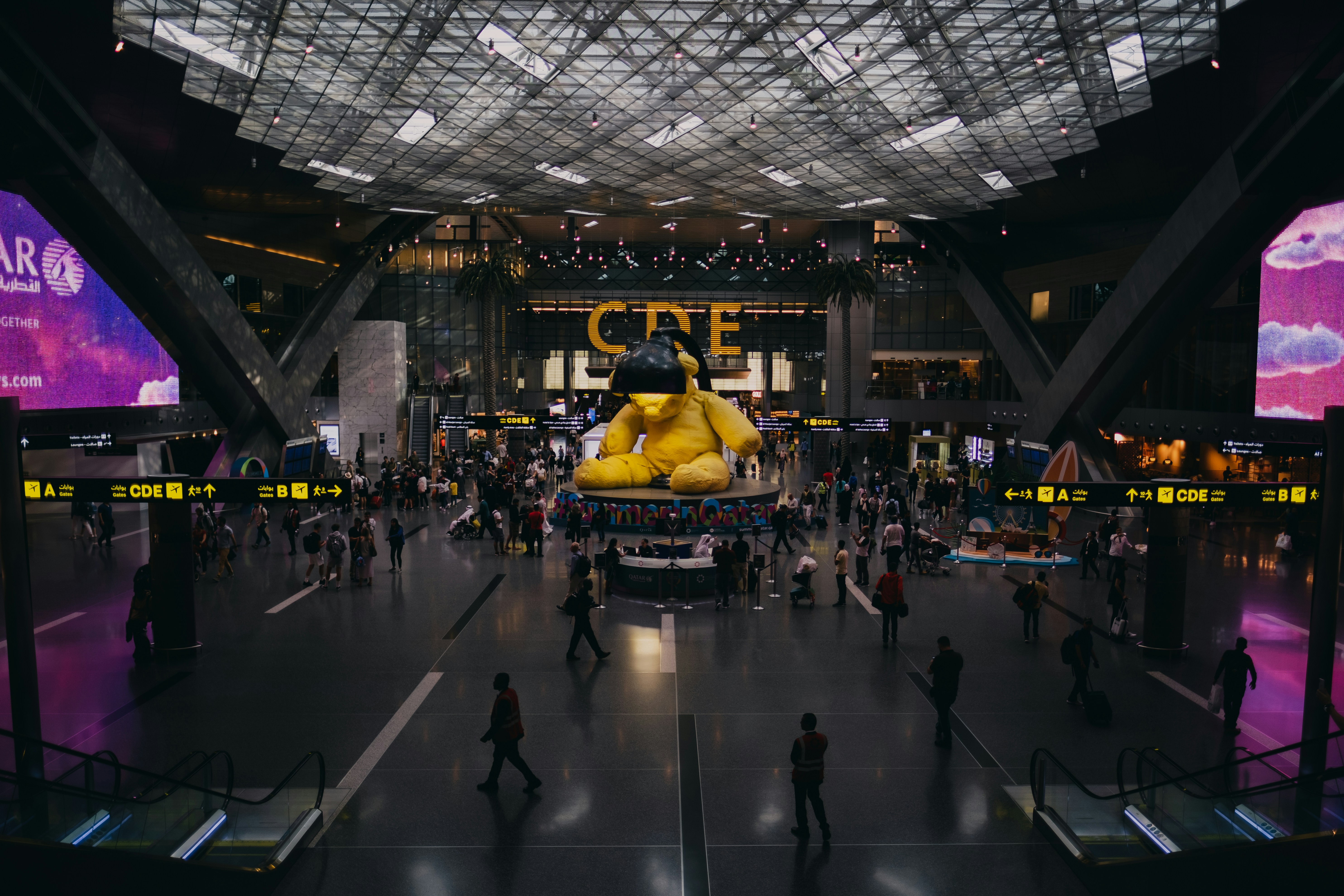 Vibrant airport terminal with iconic yellow teddy bear sculpture under a geometric glass ceiling.
