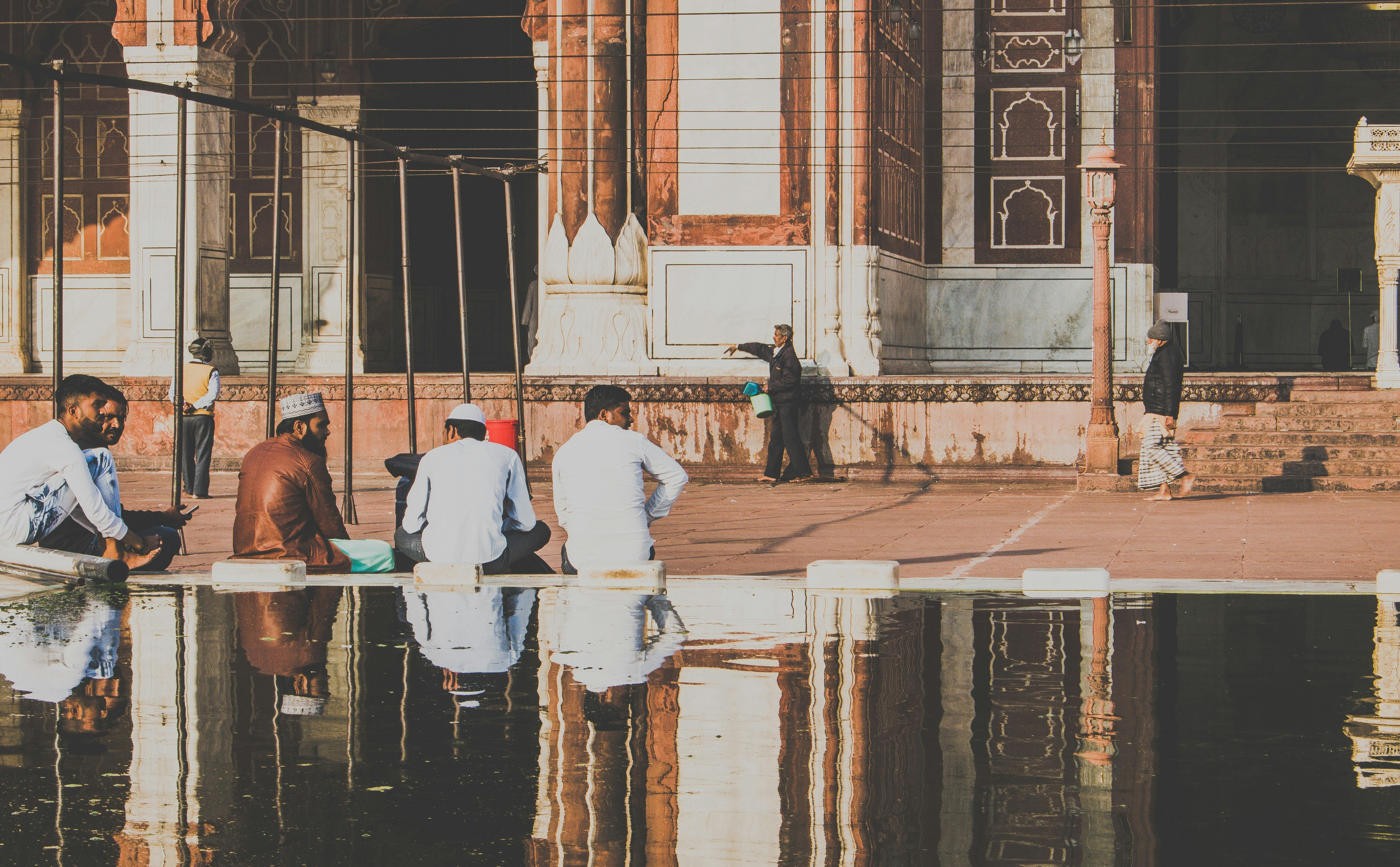 Men seated by a reflecting pool in front of a mosque, with intricate architectural details and warm sunlight.