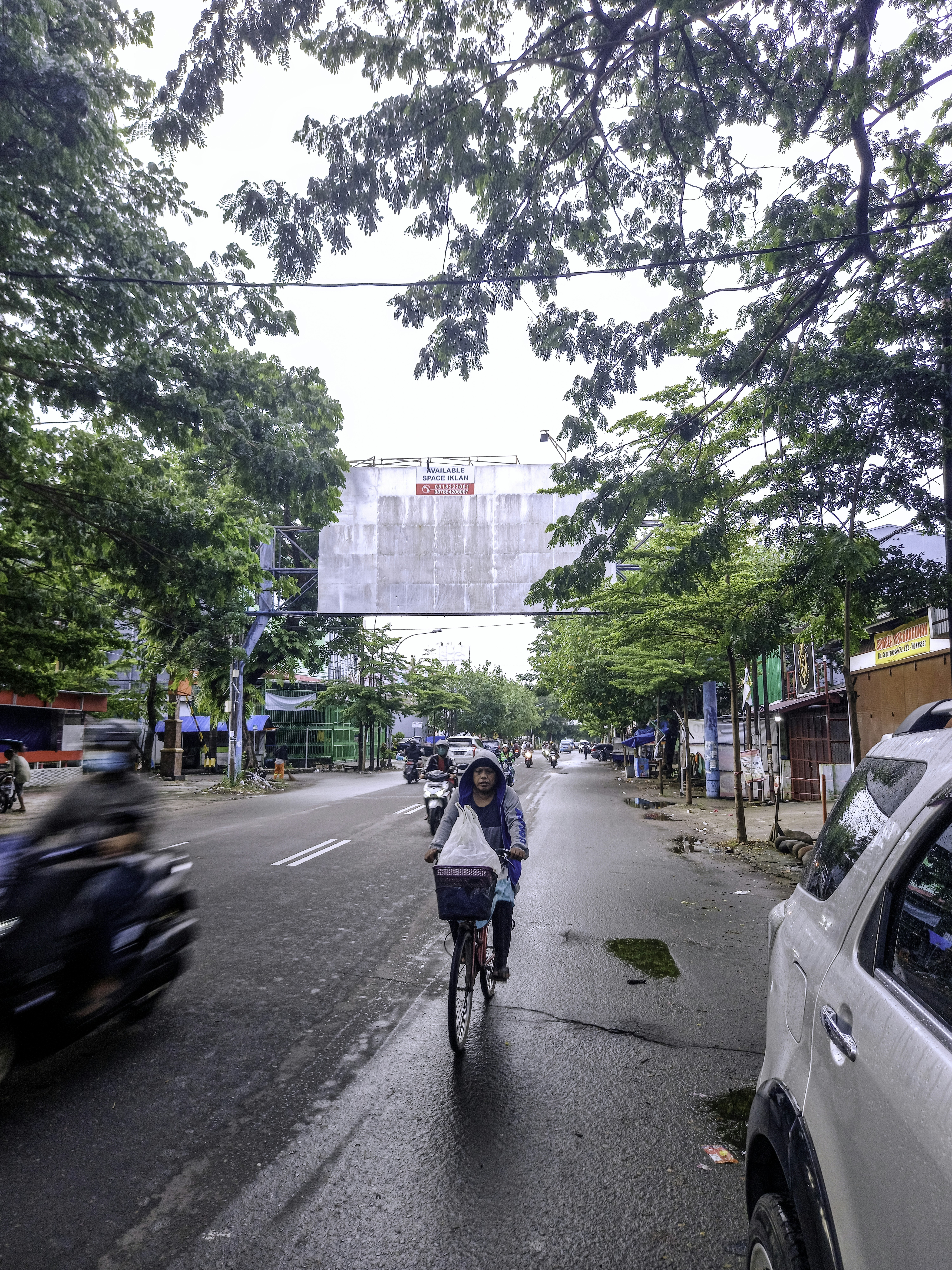 A cyclist navigates a tree-lined street, carrying a load while surrounded by urban life. The scene captures the essence of daily commuting.