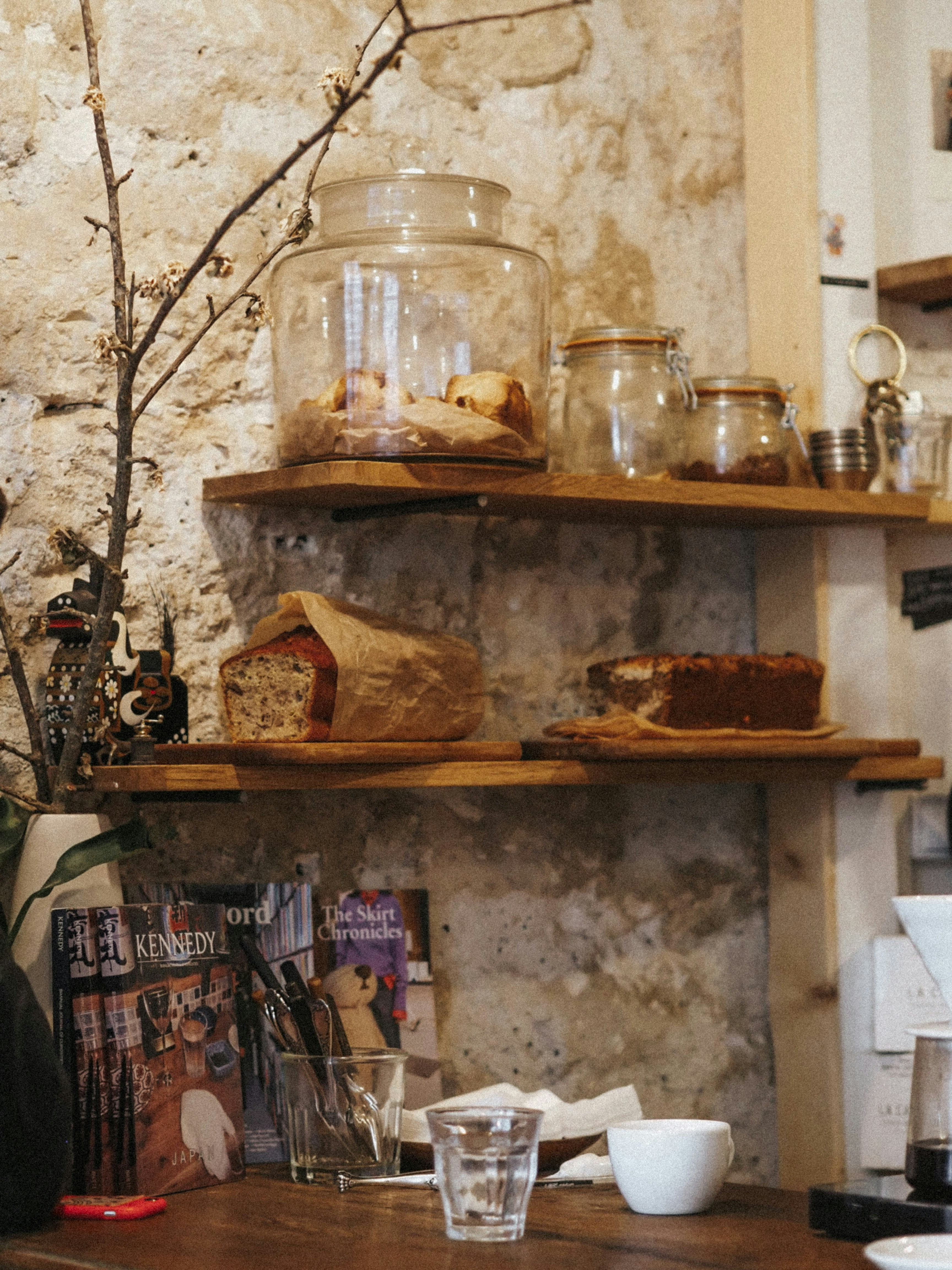 Warm wooden shelves display freshly baked goods and jars, complemented by a rustic stone wall. A branch adds a touch of nature to the inviting café atmosphere.