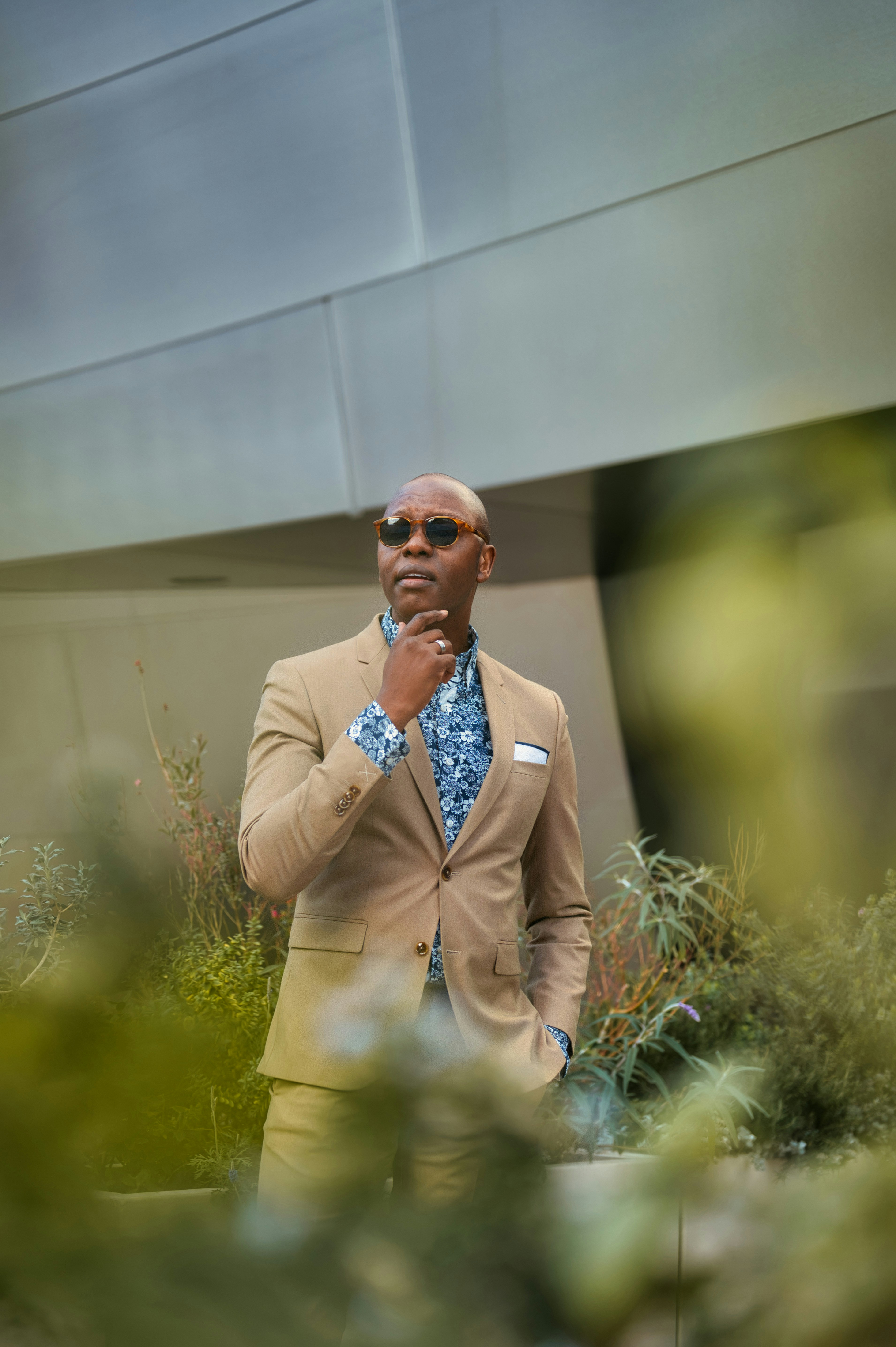 A dapper man in a tan suit and floral shirt stands thoughtfully amidst soft greenery, framed by modern architecture.