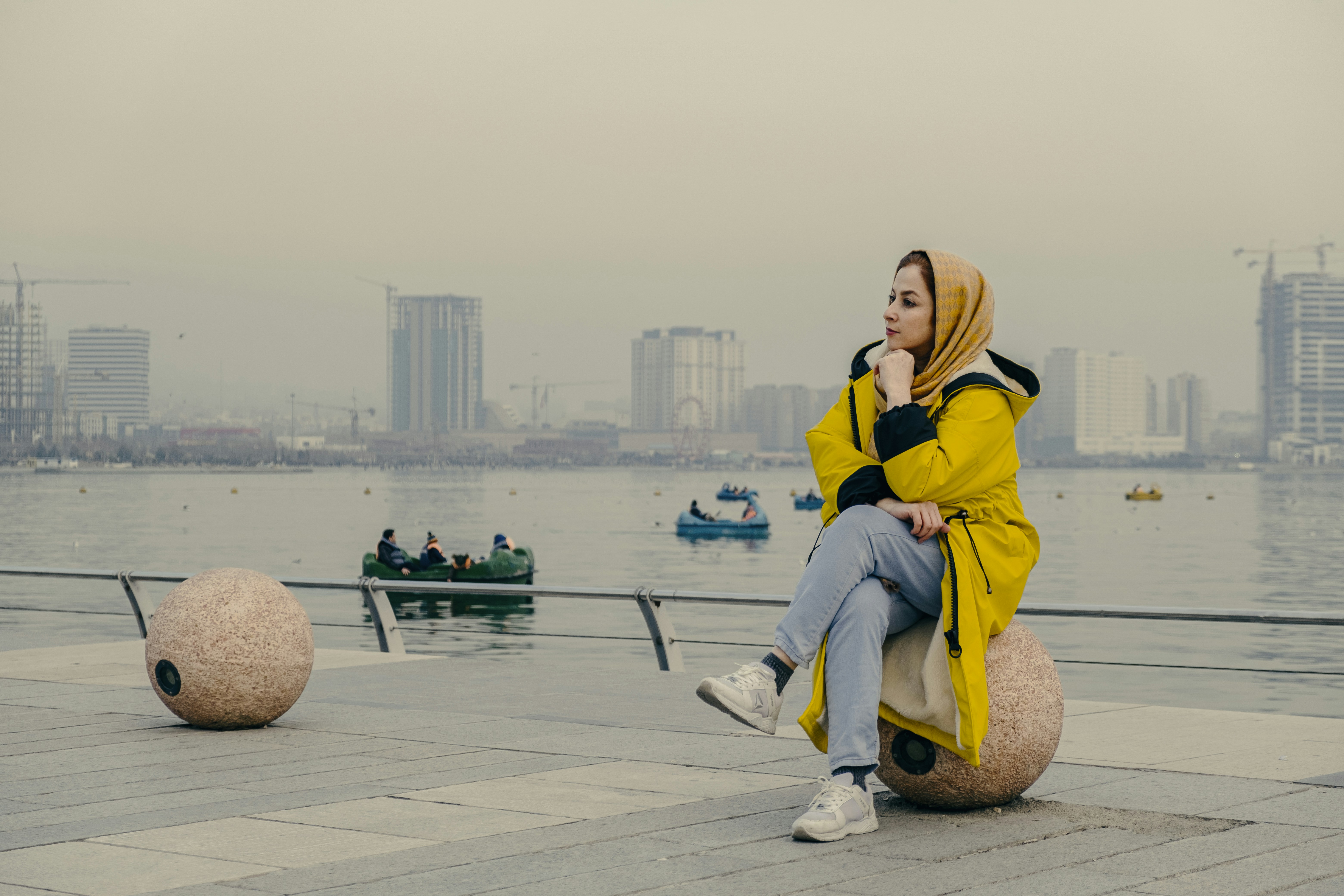 Woman in a yellow coat sitting thoughtfully on a spherical stone by the waterfront, with boats and city skyline in the background.