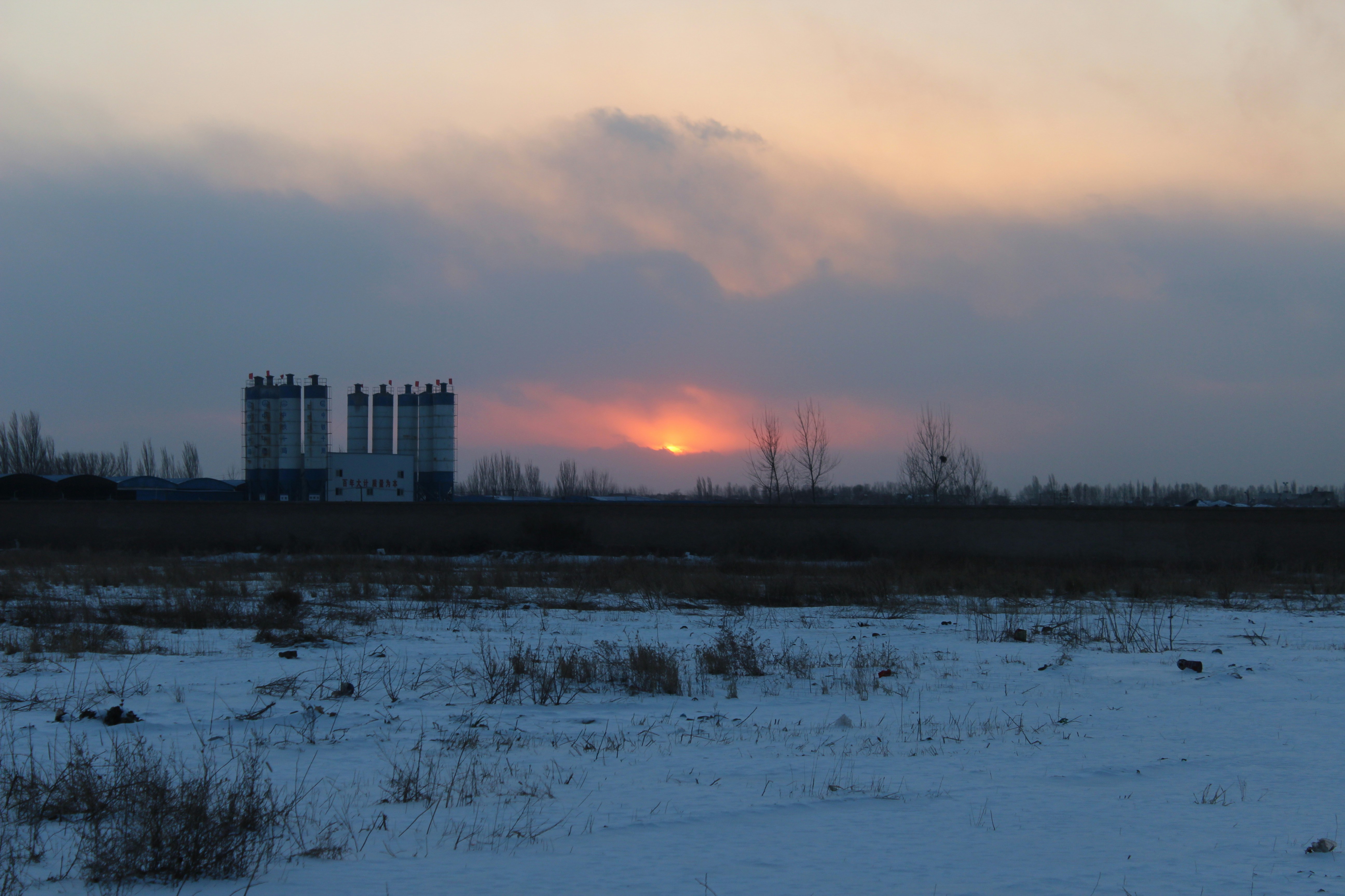 Industrial buildings stand silhouetted against a vibrant winter sunset, casting a stark contrast with the snowy landscape.