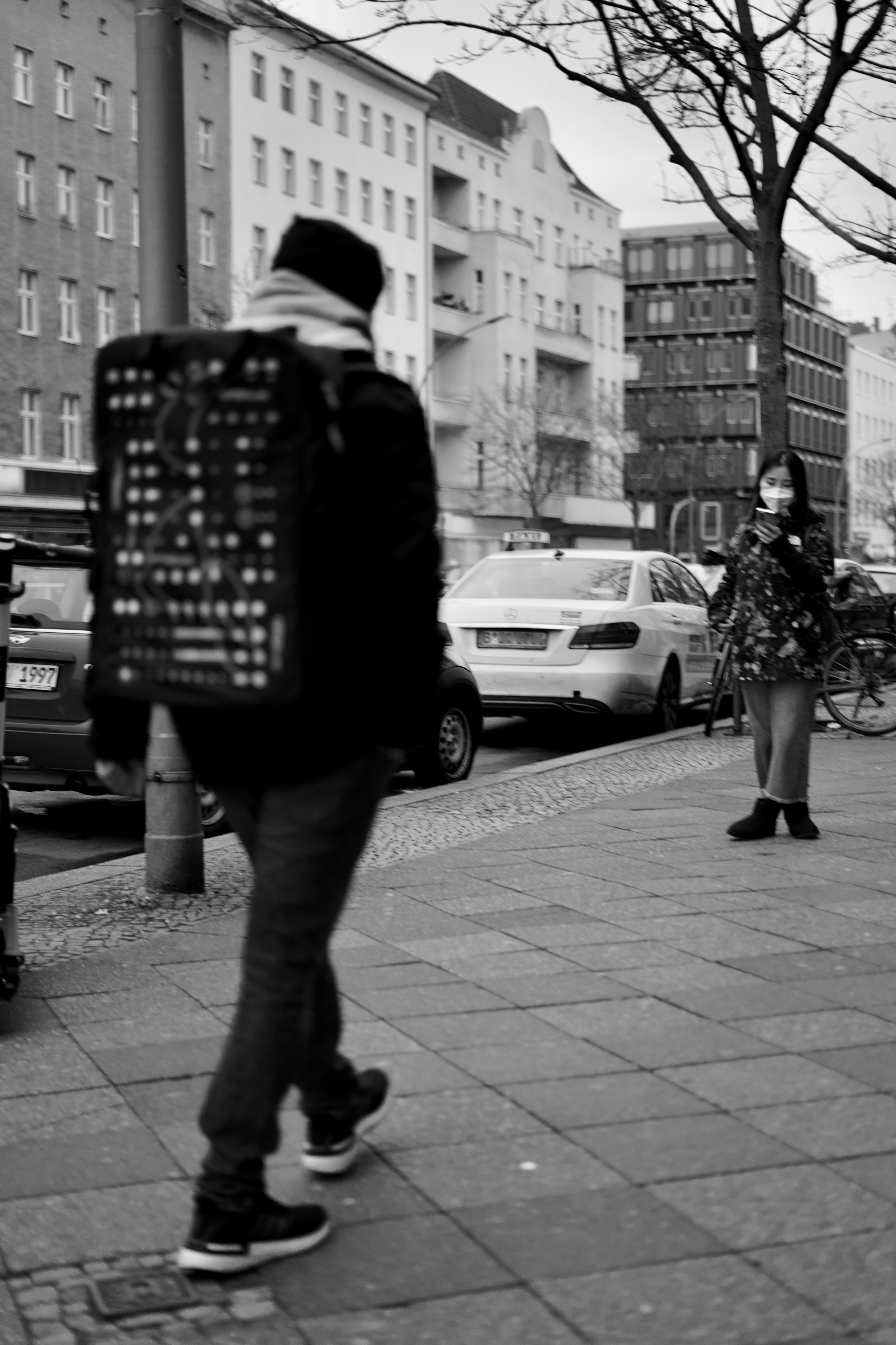 A man walks past a woman engrossed in her phone on a bustling city street, capturing the essence of urban life. The scene is set against a backdrop of modern architecture.