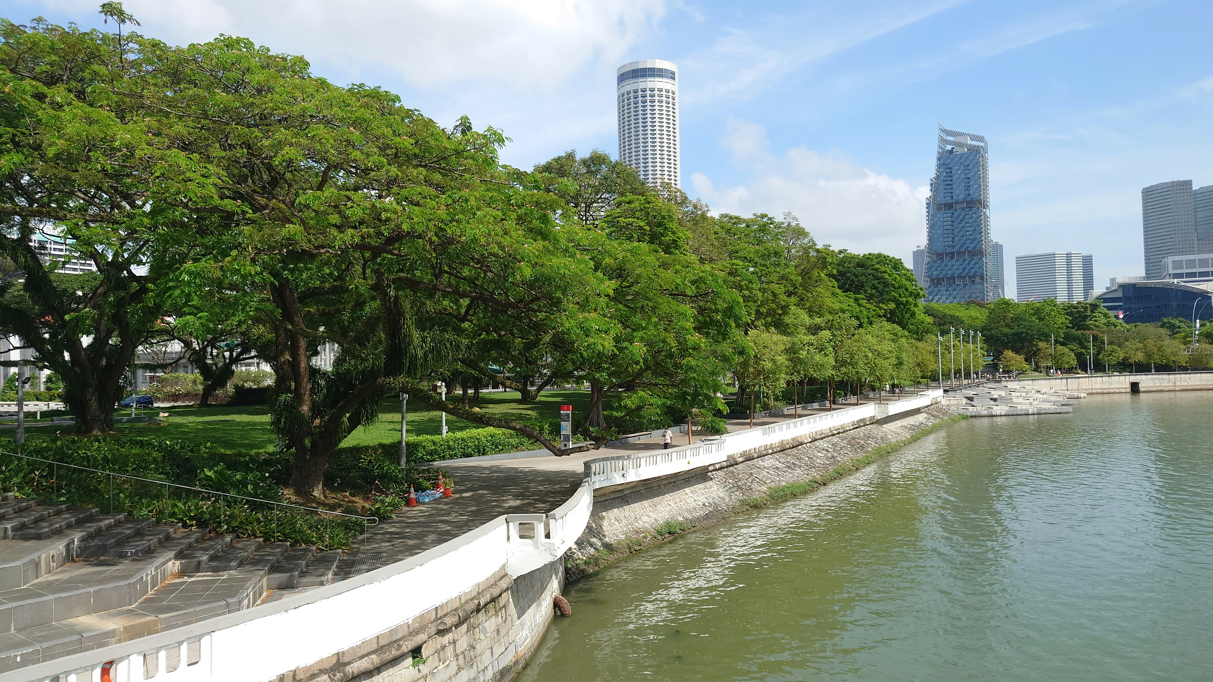 Lush green trees line a riverbank with modern skyscrapers in the background.