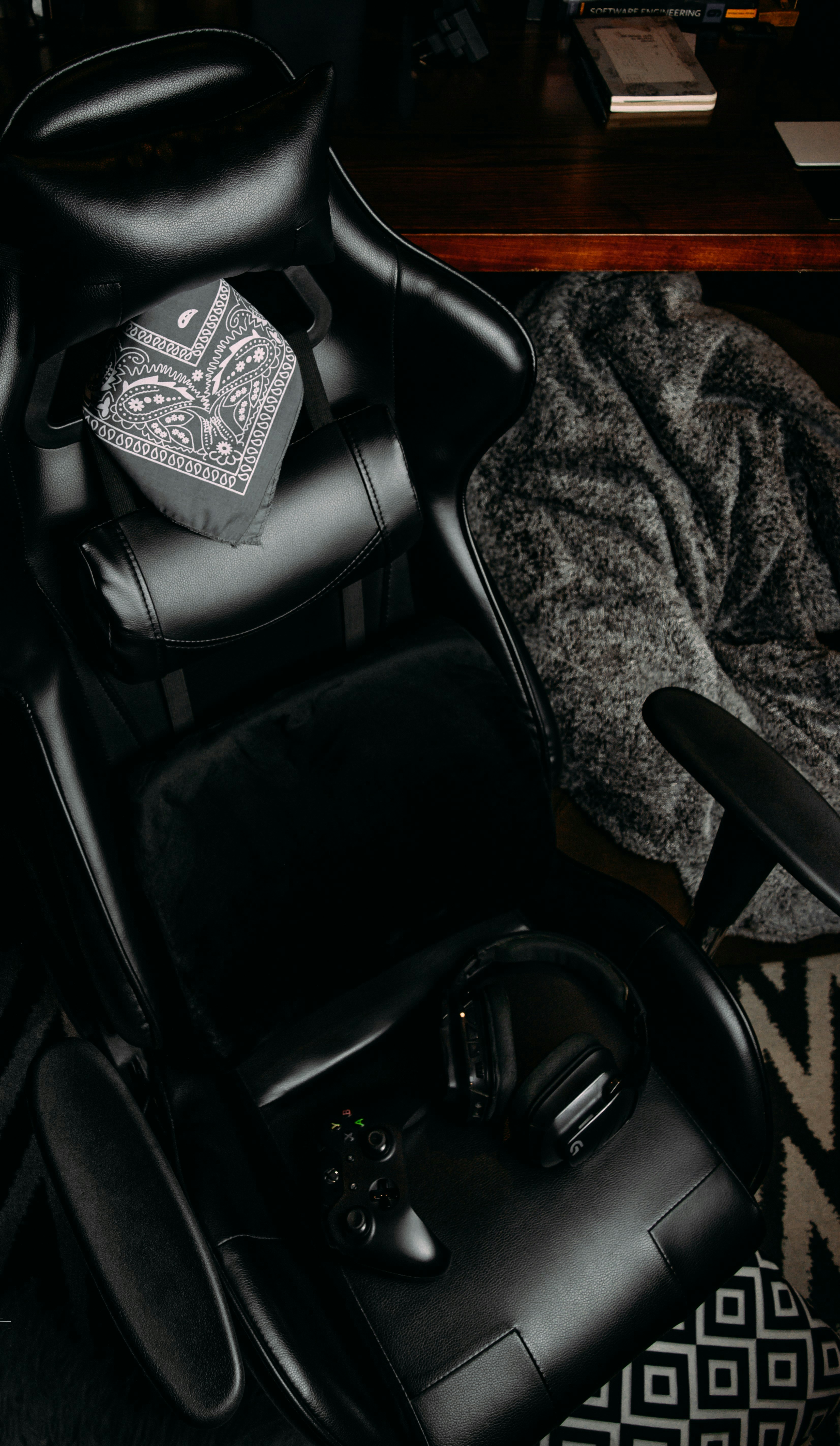 A sleek black gaming chair adorned with a patterned bandana, accompanied by a gaming headset and controller, set against a textured blanket and patterned rug.
