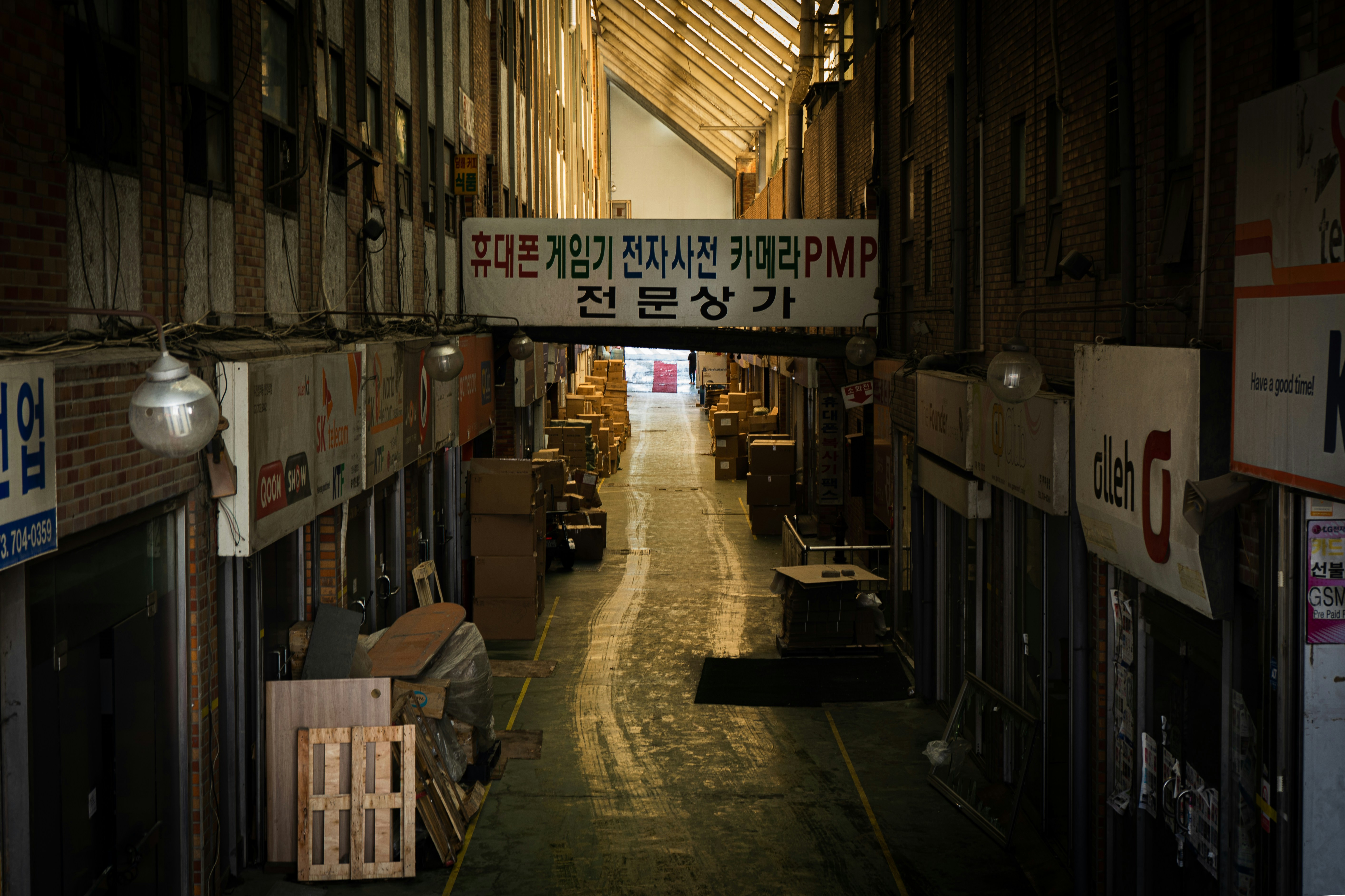 Dimly lit corridor of a deserted market, lined with stacked boxes and illuminated by overhead lights. A large sign hangs above, hinting at the space's former vibrancy.