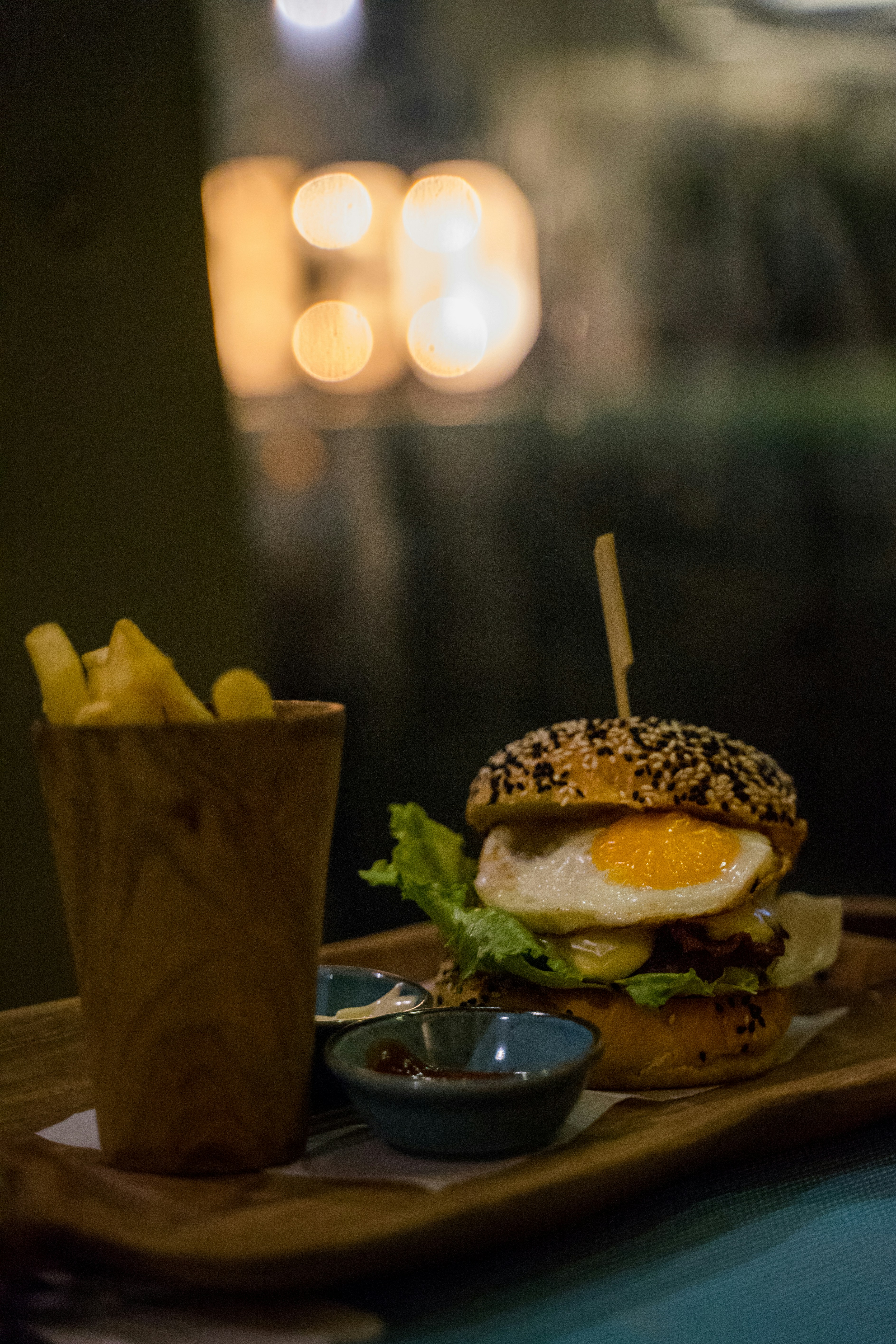 A beautifully arranged gourmet burger topped with a fried egg, accompanied by crispy fries and dipping sauces. The setting features a softly blurred background of ambient lights.