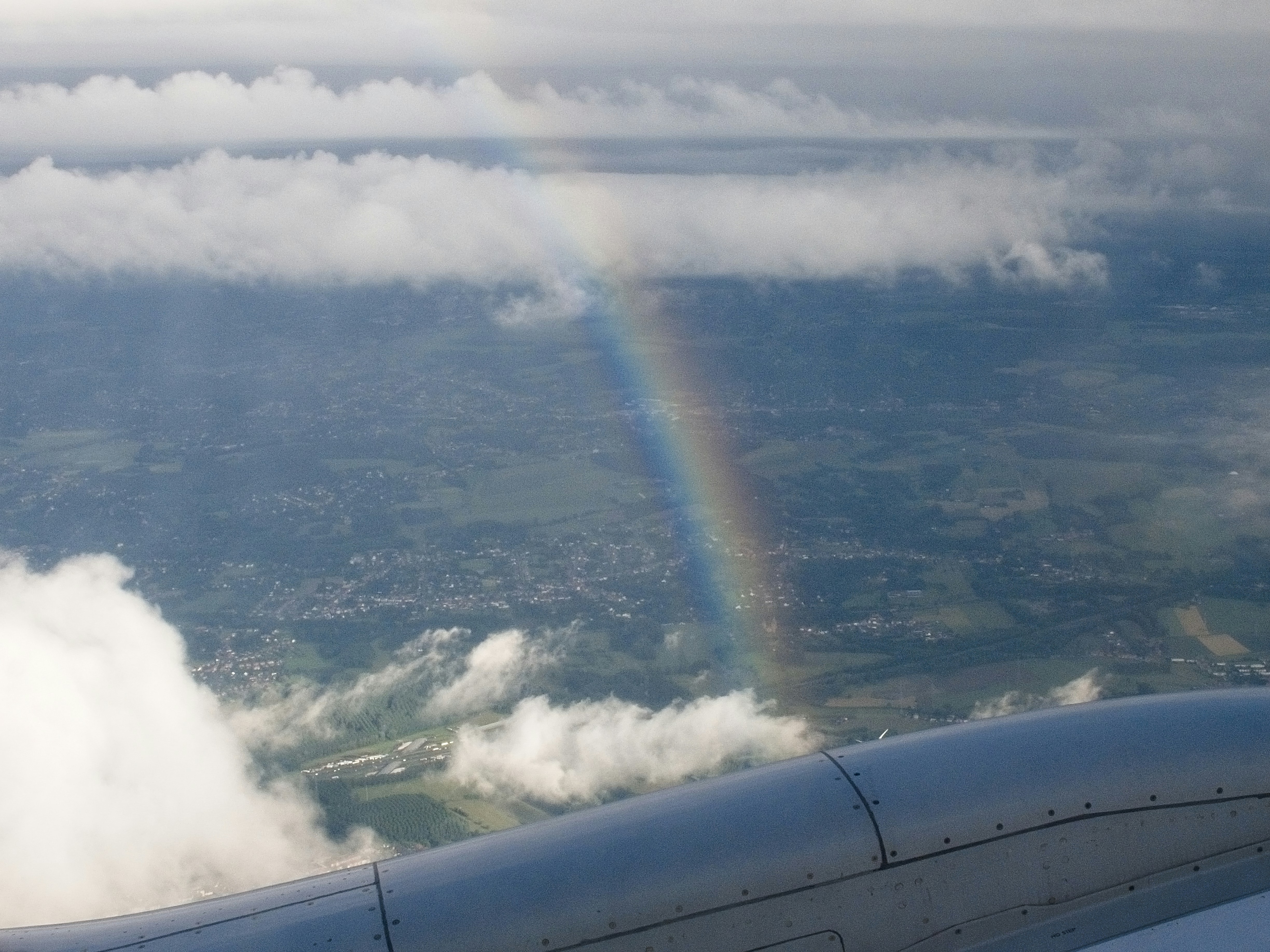 Vibrant rainbow arching over a lush landscape, viewed from an airplane window amidst fluffy clouds. The metallic wing adds a contrasting element to the scene.