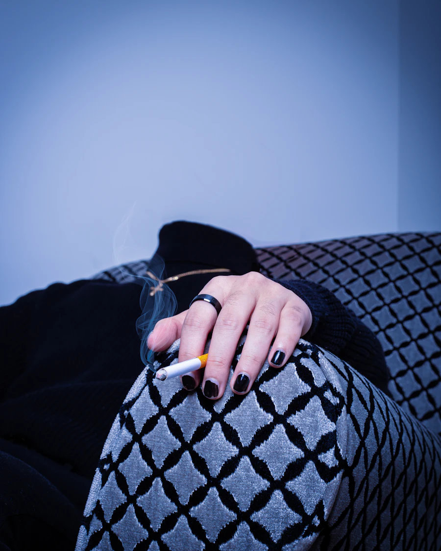 A hand with black nail polish and a ring rests on patterned fabric while holding a lit cigarette with smoke curling upward. The scene is lit in cool tones against a plain wall.