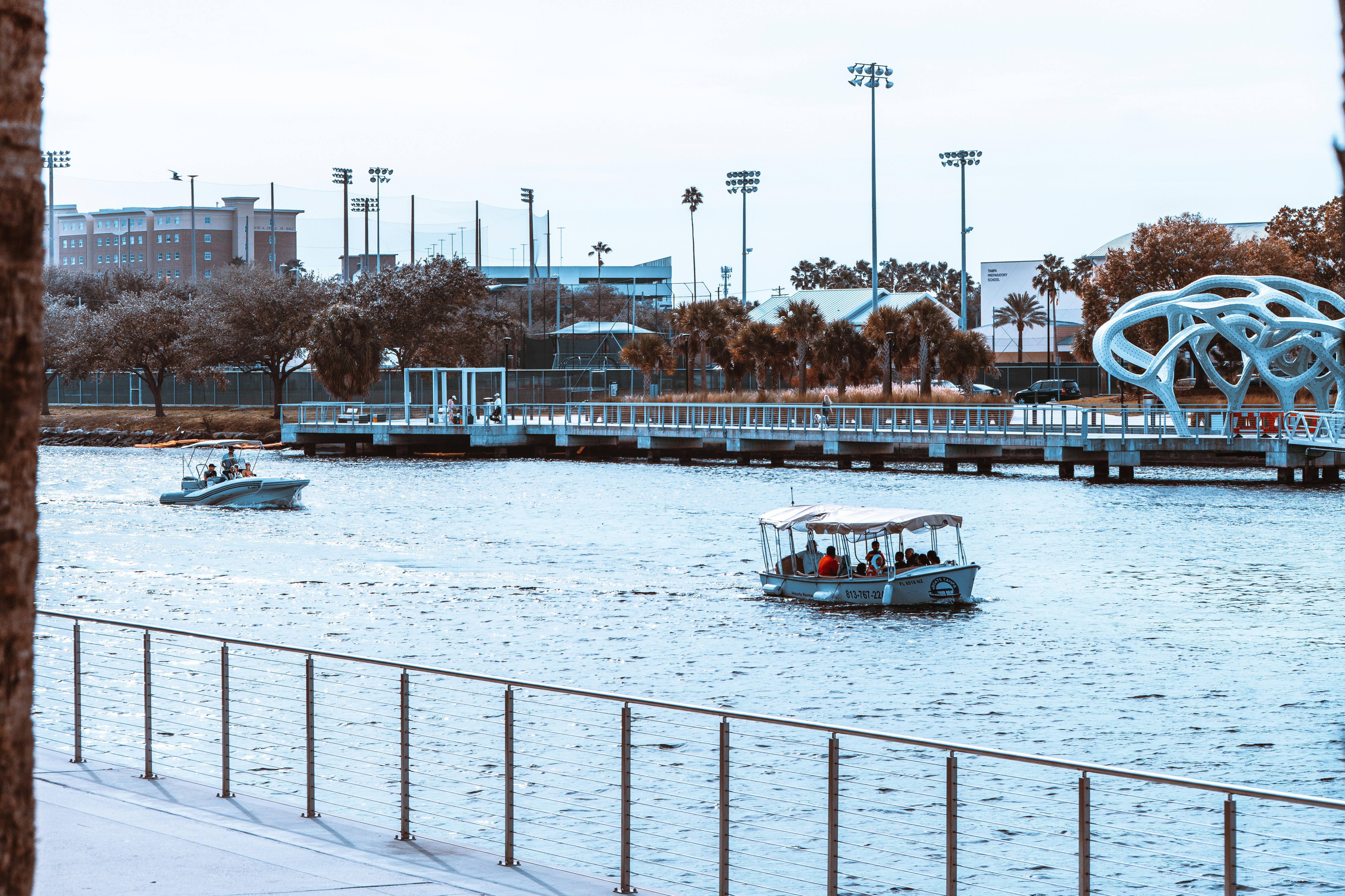 Leisure boats navigate a city river near a modern pier, with urban structures and palm trees in the background.