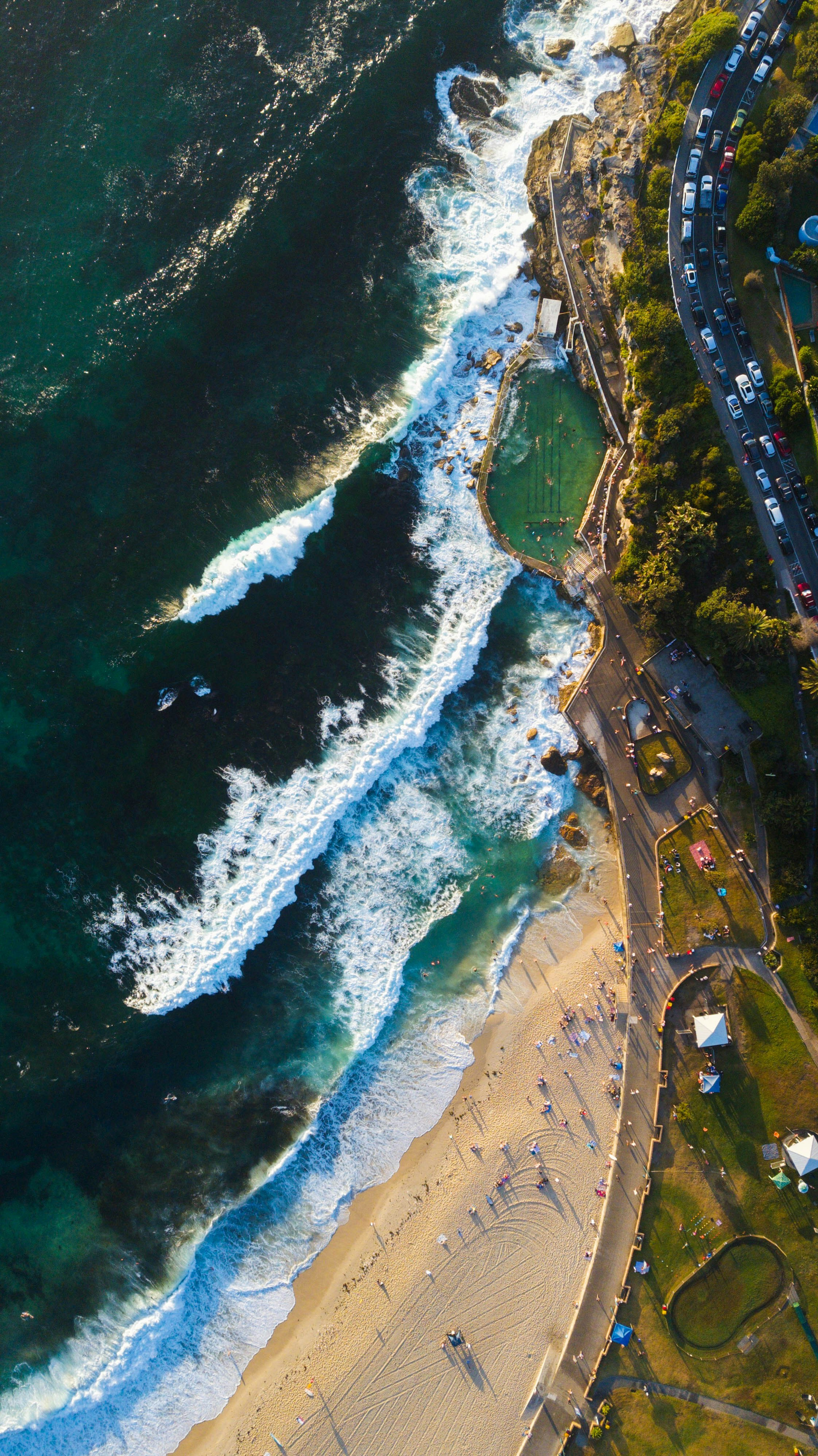 Aerial view of a beachside scene featuring crashing waves, sandy shore, and a winding road lined with cars. The vibrant colors of the ocean contrast beautifully with the golden sand.