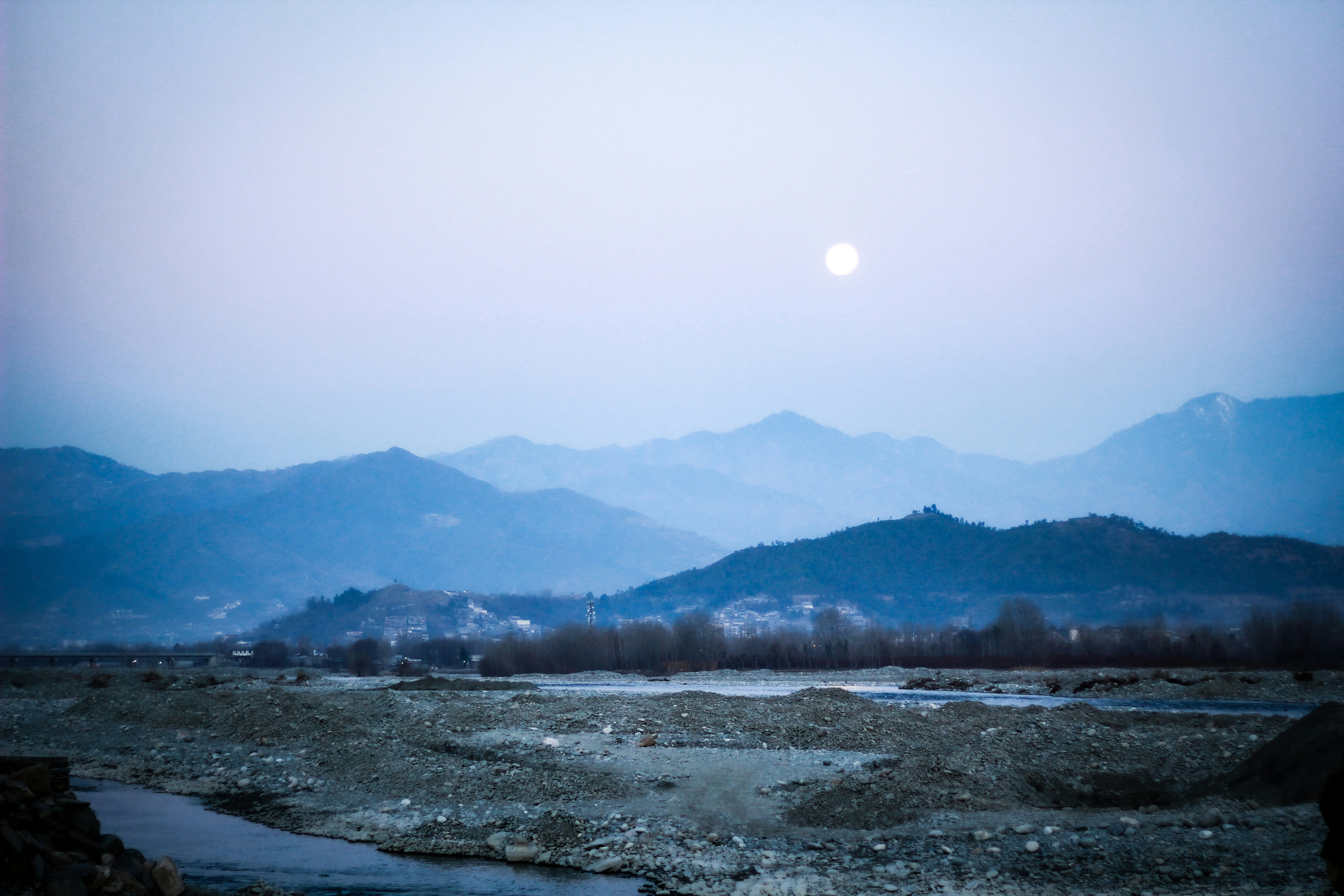 A tranquil landscape featuring distant mountains under a glowing full moon, with a calm riverbed in the foreground. The soft hues of twilight create a serene atmosphere.