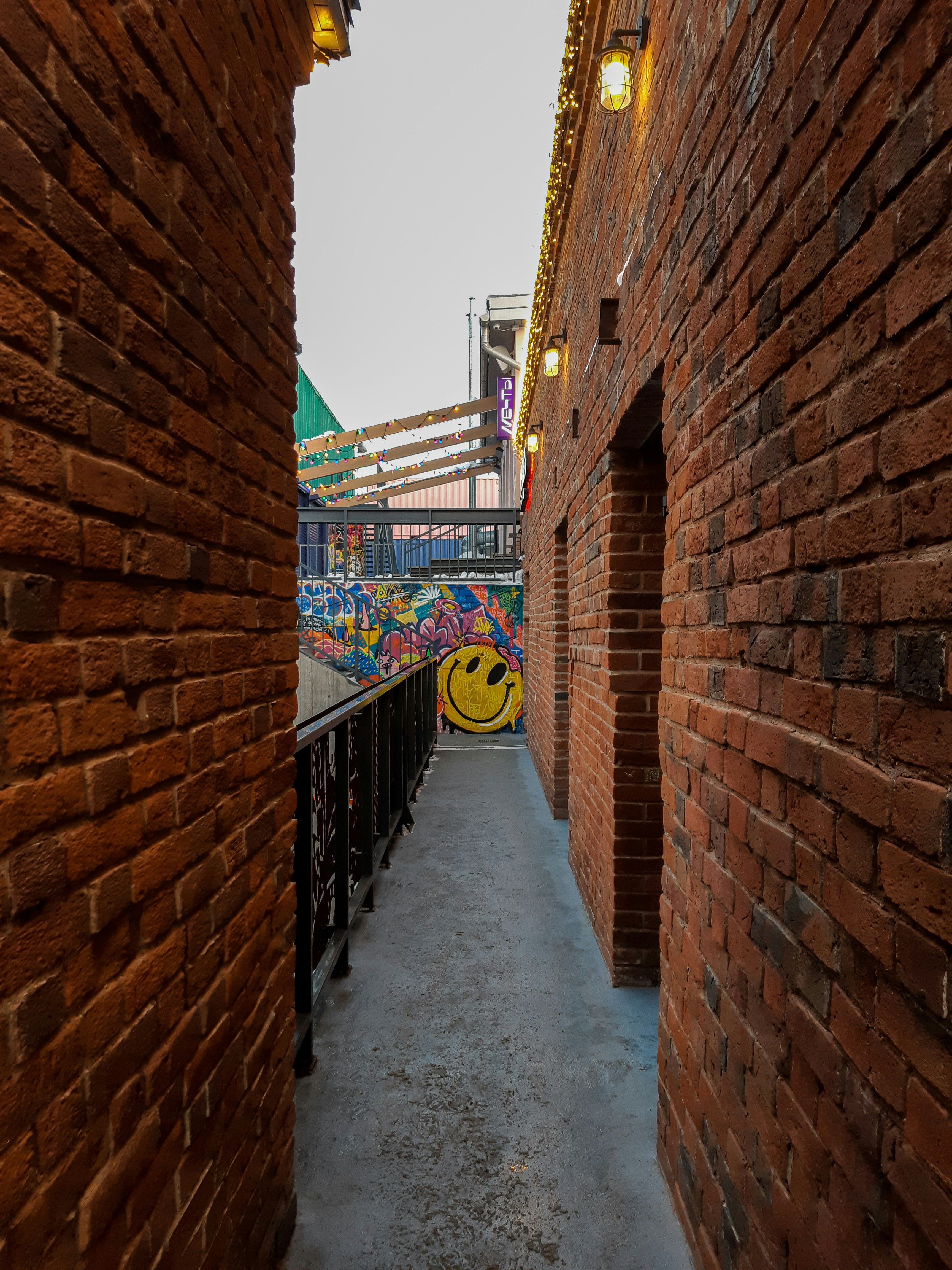 Narrow corridor flanked by rustic brick walls, leading to a colorful mural featuring a smiling face. The space is illuminated by warm lights above.