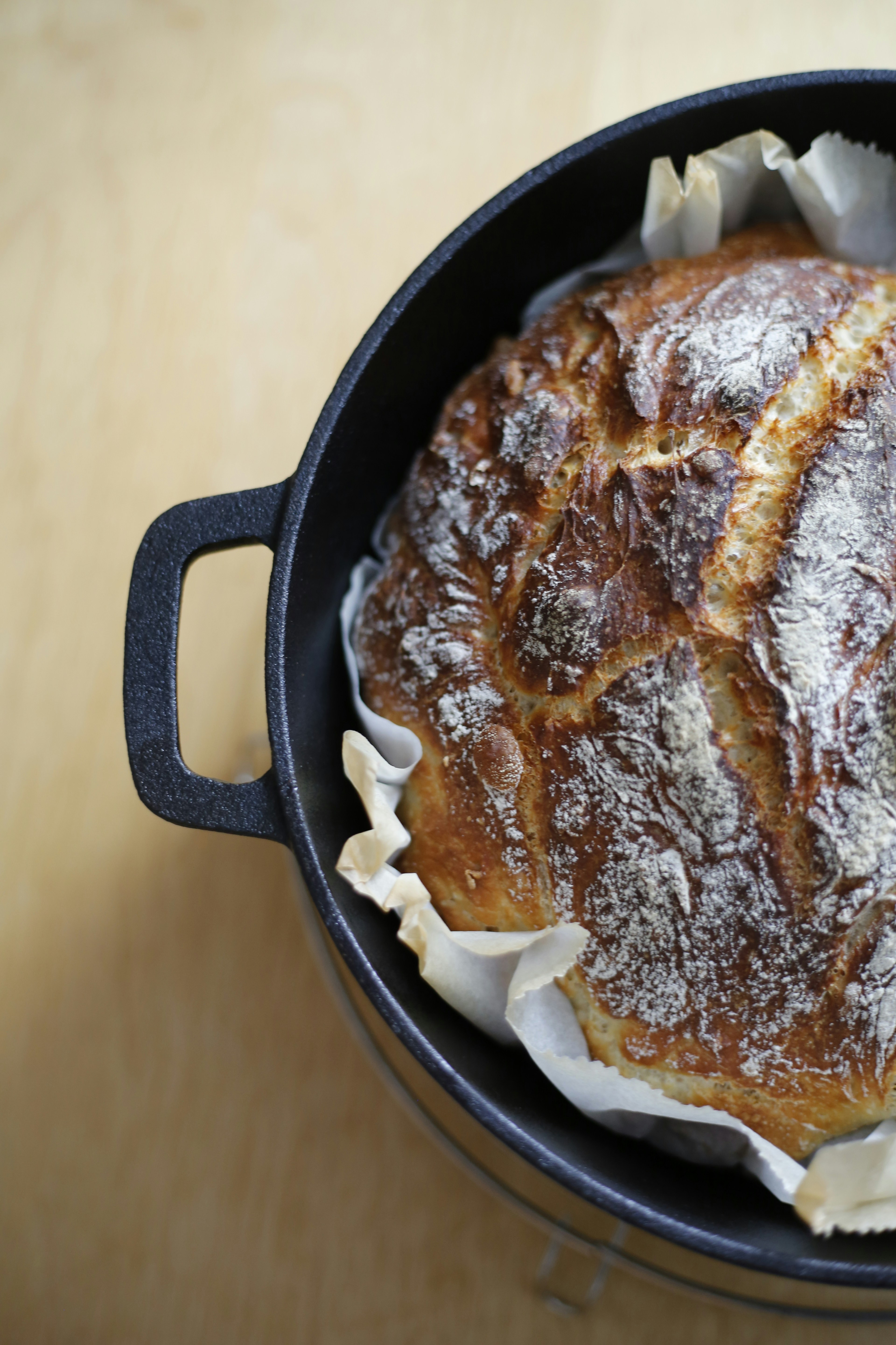 Golden-brown artisan bread resting in a cast iron pot, showcasing a beautifully cracked crust and parchment paper lining. 