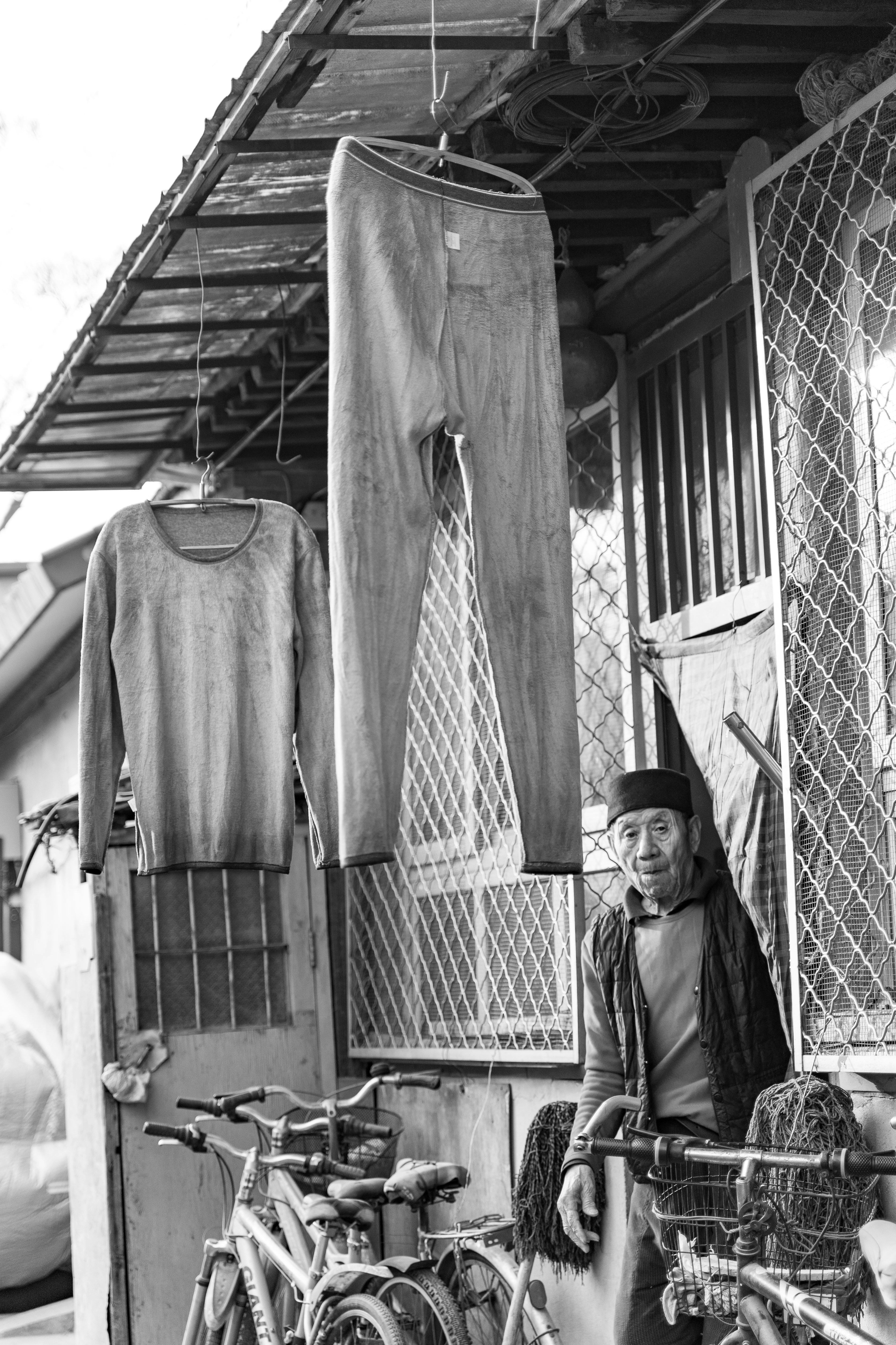 An elderly man stands near bicycles, gazing thoughtfully at hanging garments in a rustic setting. The monochrome palette emphasizes the textures and details of the scene.