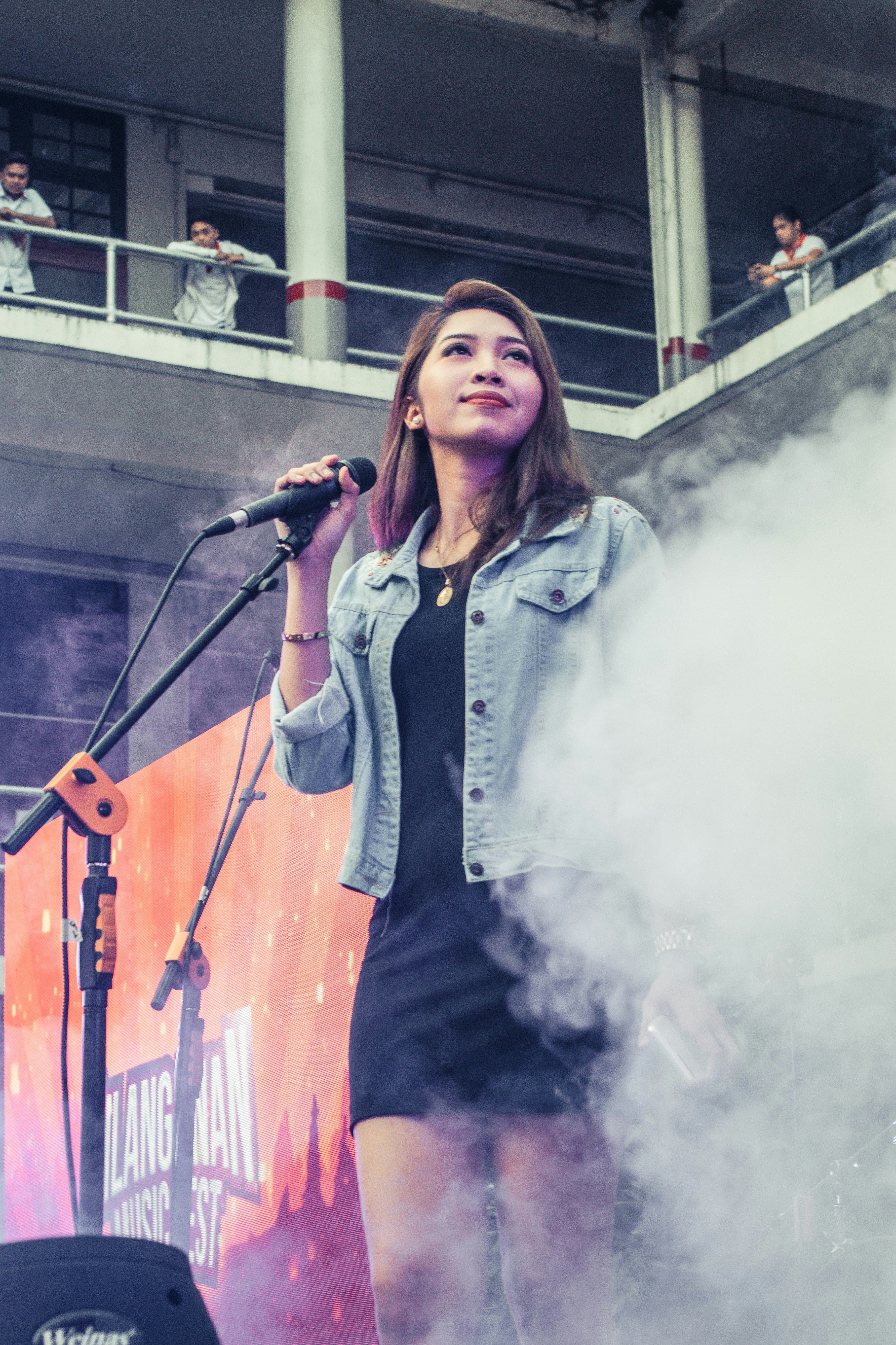 A performer stands confidently on stage, microphone in hand, with a backdrop of vibrant lights and a smoky atmosphere.