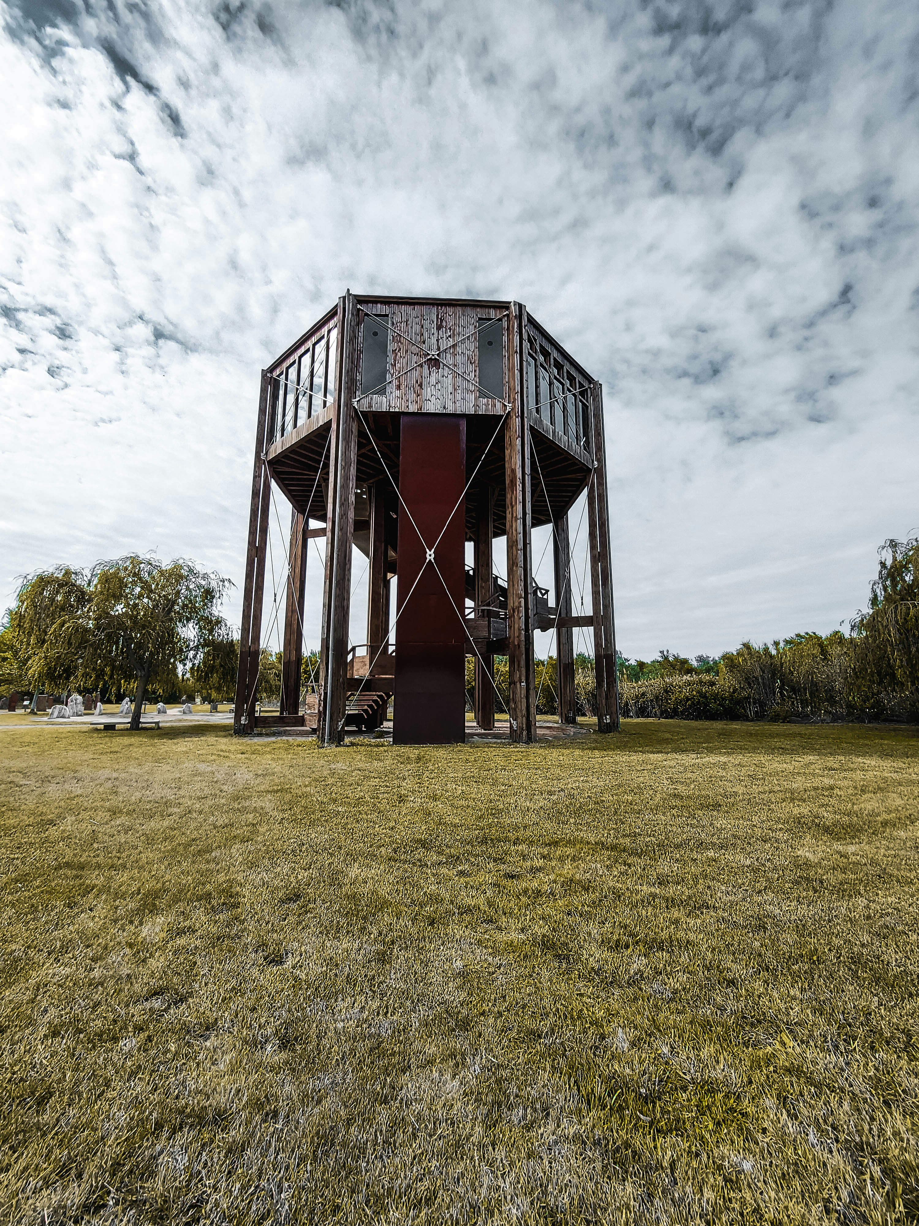 Weathered wooden observation tower rises over a sunlit meadow beneath a dramatic, cloud-filled sky.