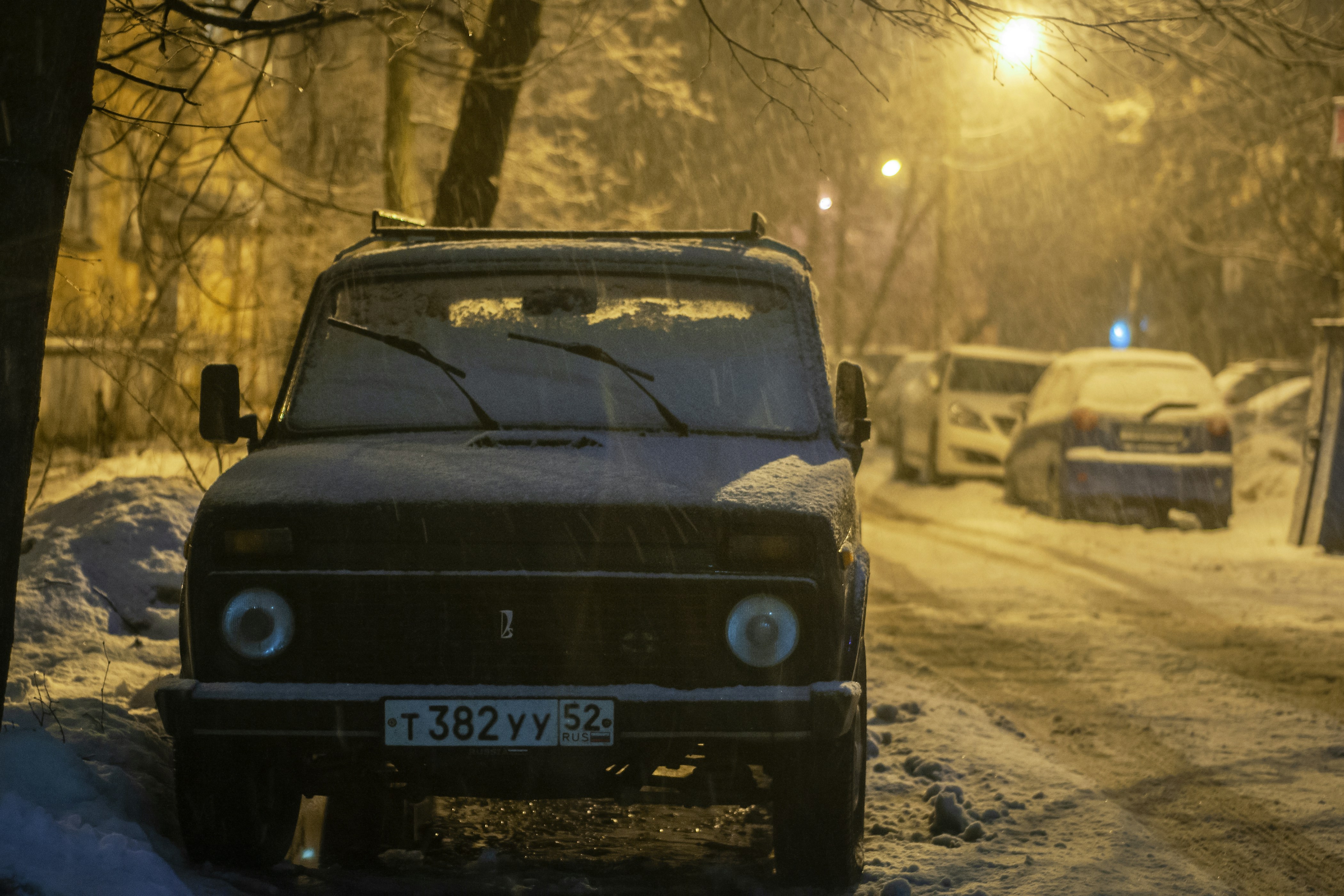 A vintage car blanketed in snow, parked on a quiet street illuminated by warm streetlights during a snowfall.