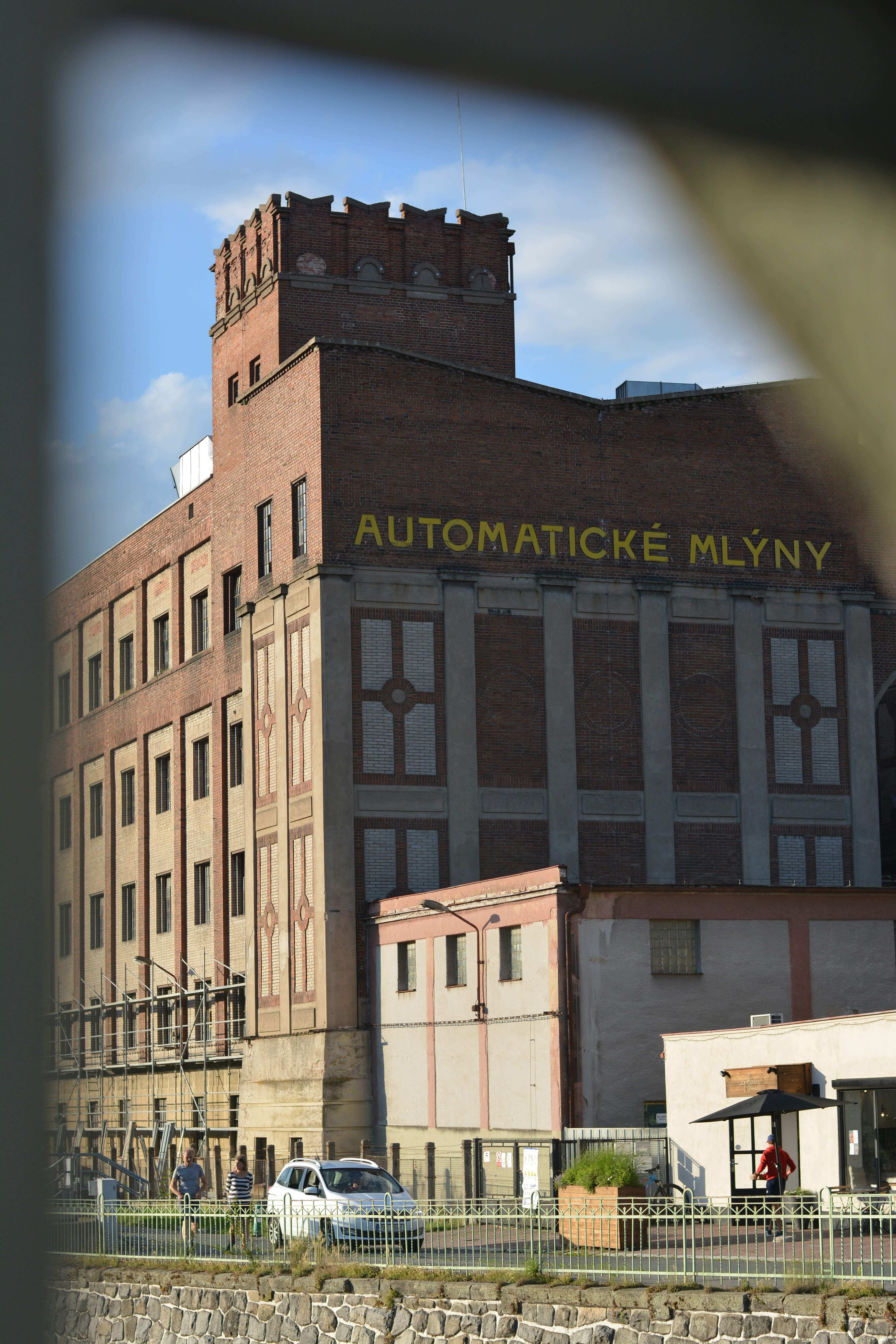 Brick industrial facade of the Automatické Mlýny building seen through a framing window, with a street, fence, and a white car.