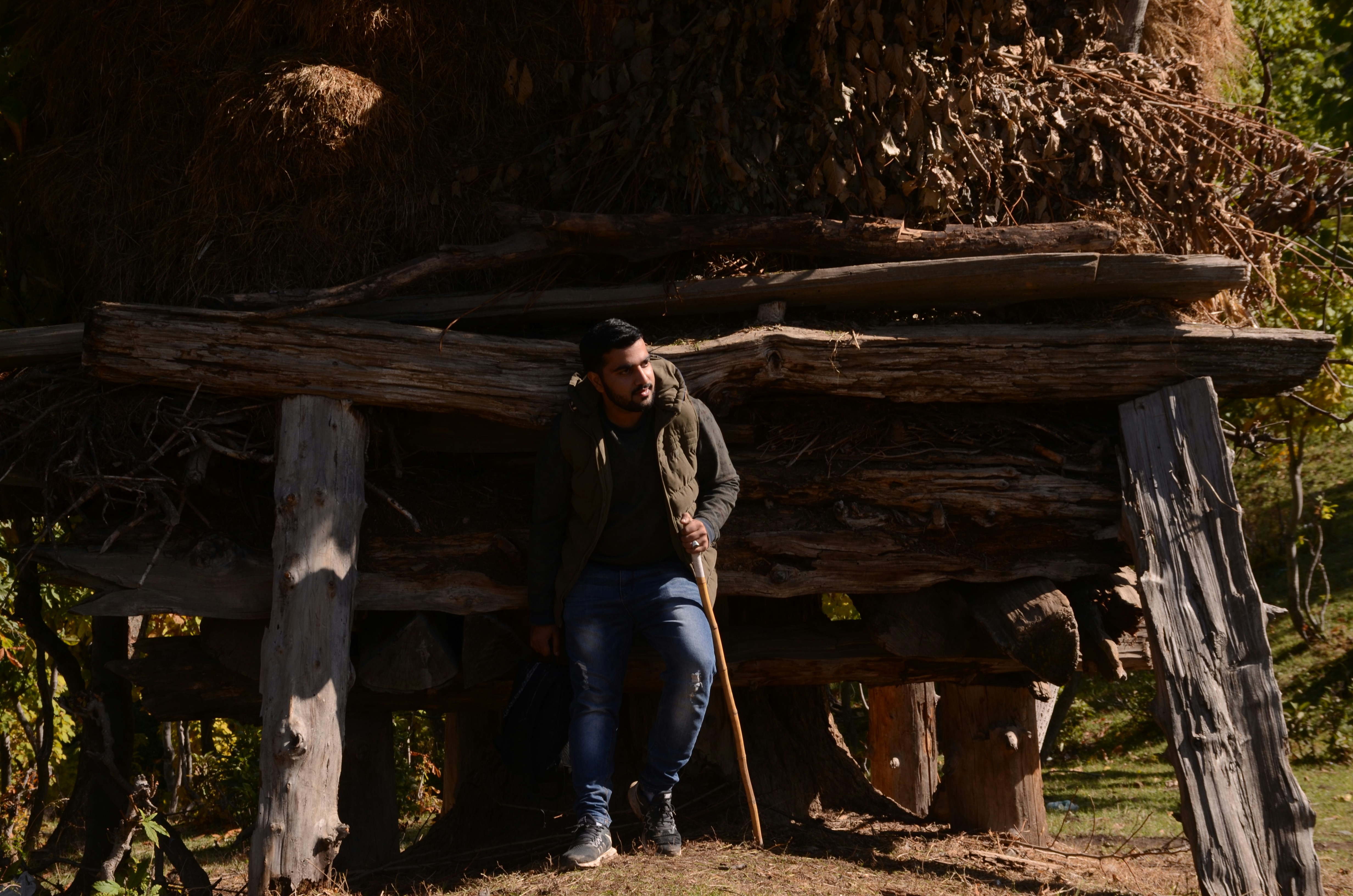 A man sitting against a rustic wooden structure, holding a walking stick amidst a serene natural setting.