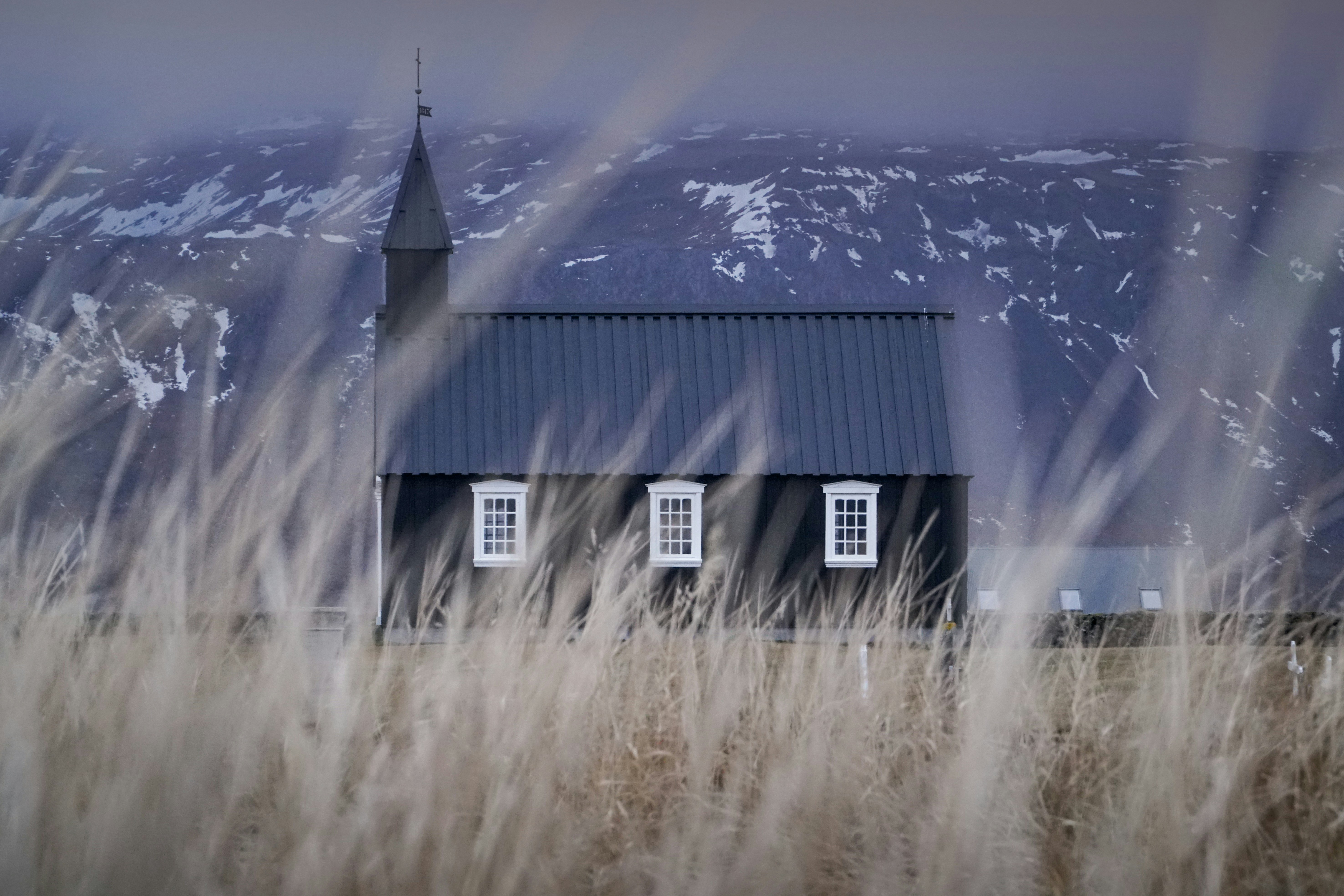 Black wooden chapel stands against a backdrop of snow-capped mountains, framed by tall grass swaying in the wind.