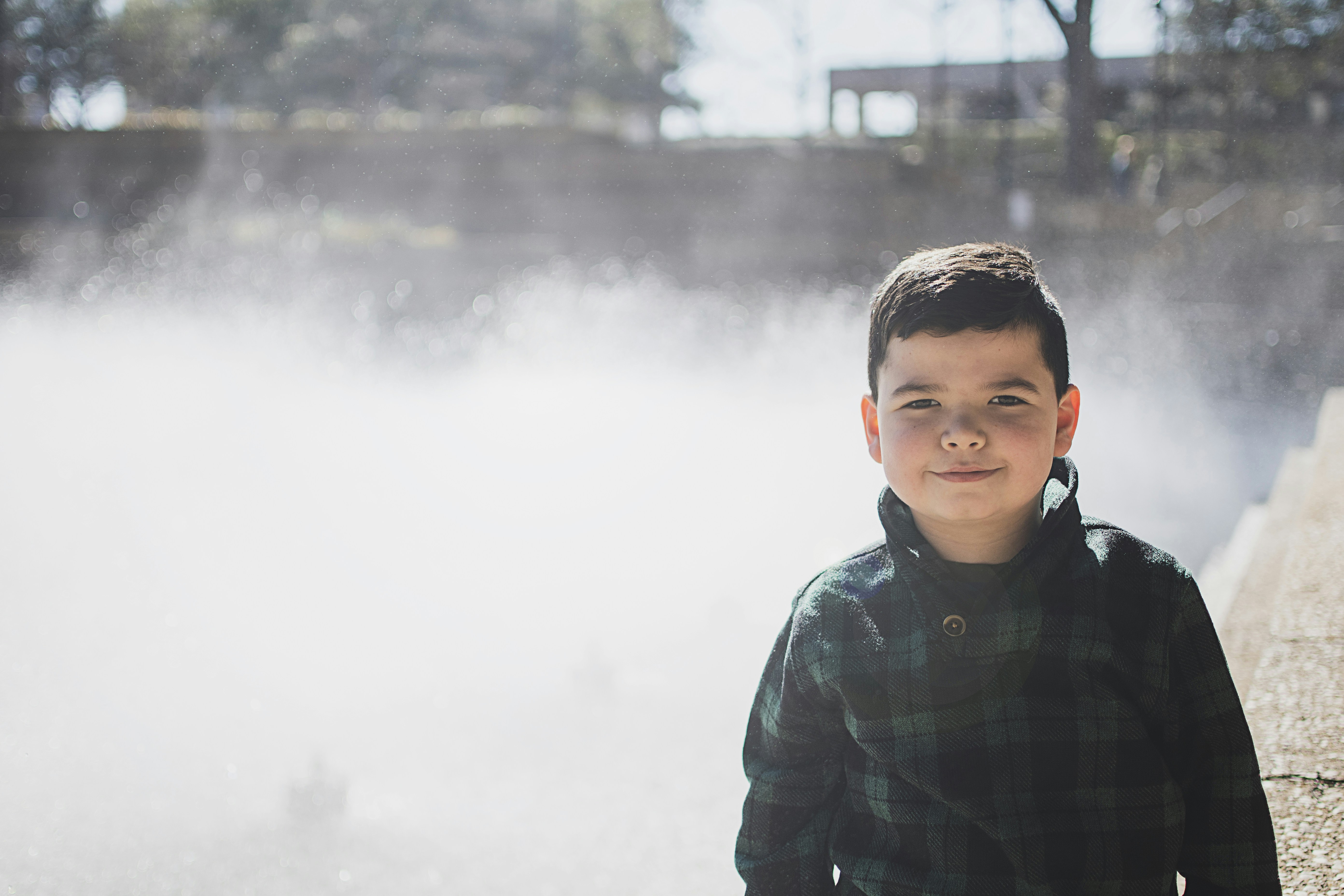 Young boy standing confidently by a misty riverside on a sunny day.