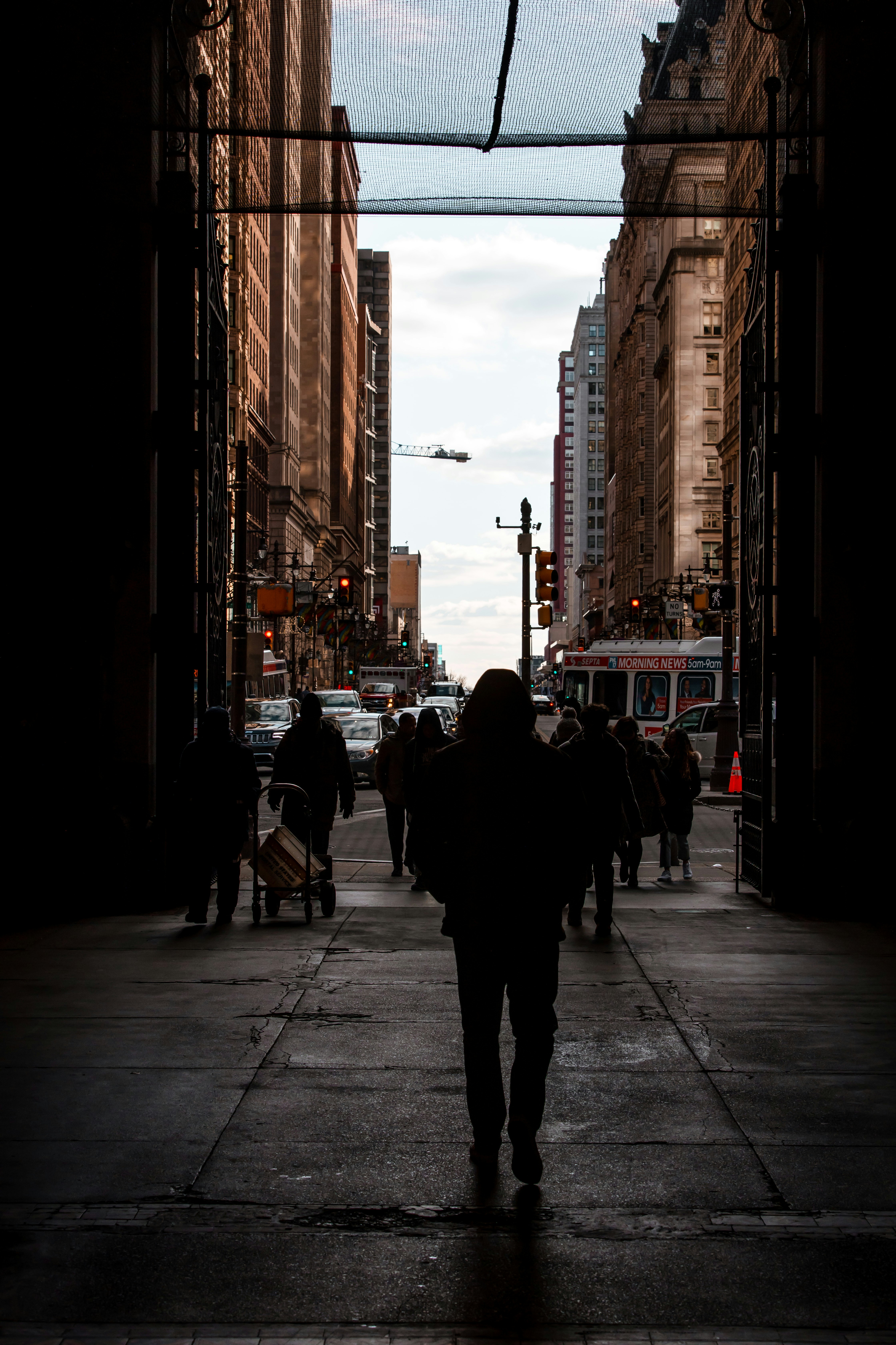 Silhouetted figures walk through a bustling urban passageway, framed by towering buildings and a hint of blue sky above. Streetlights and traffic add a dynamic backdrop.