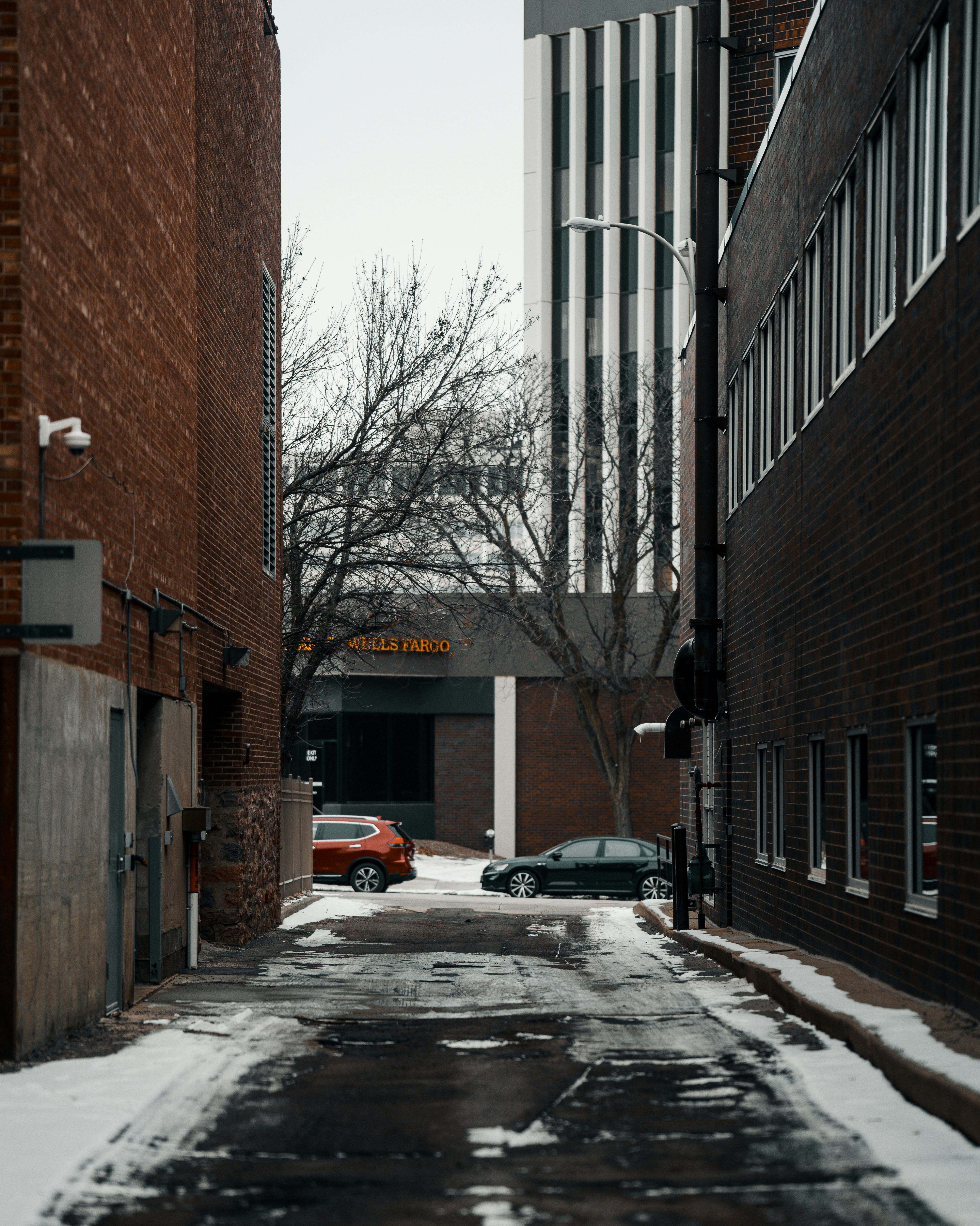 Narrow alleyway framed by brick buildings, leading to a glimpse of a street with vehicles and trees. Snow covers the ground, hinting at winter's chill.