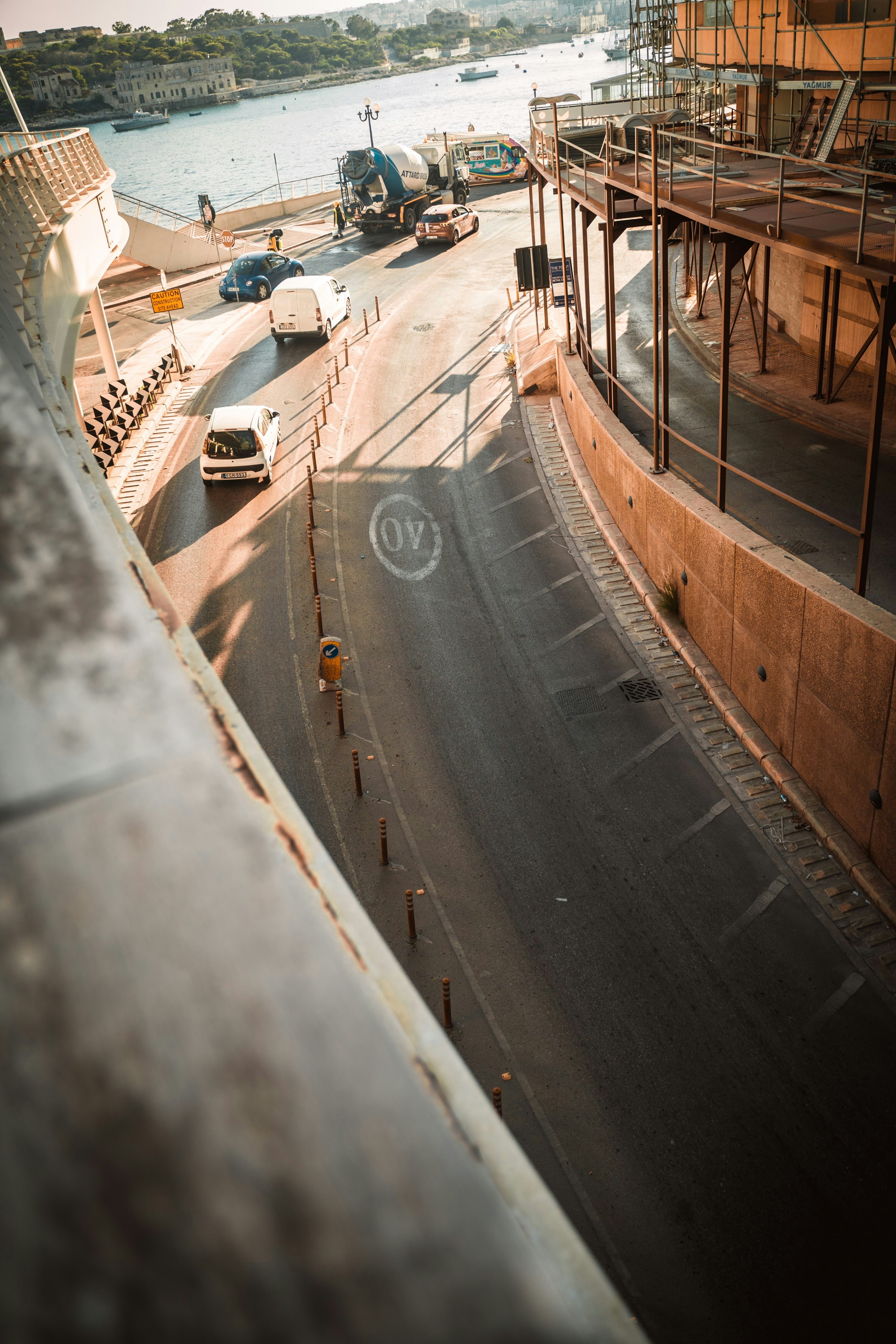 Vehicles navigating a winding road beside a waterfront, with scaffolding and urban architecture in view.