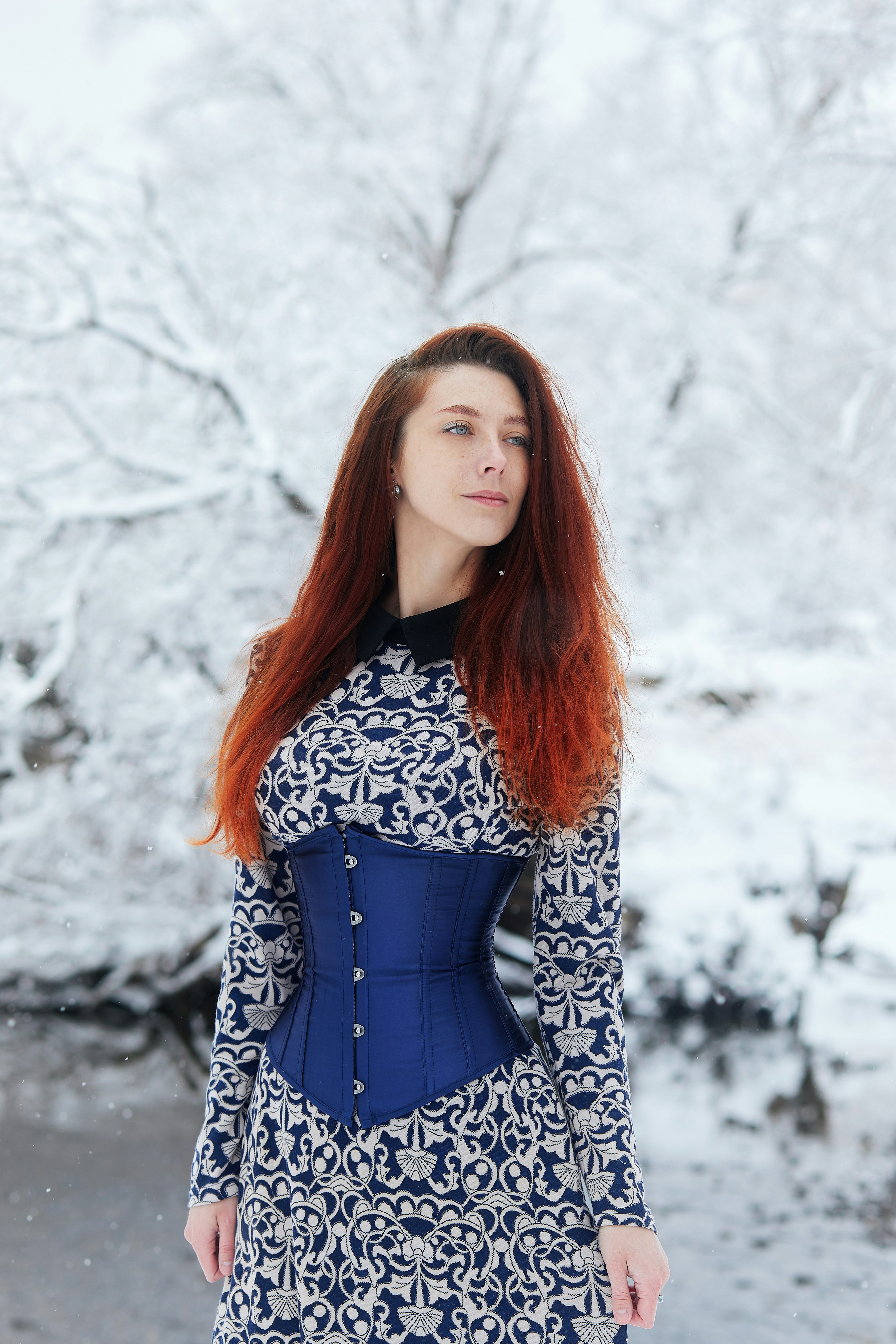A spectacular winter portrait with a beautiful red-haired girl in a long dress and blue corset. Charming young woman posing in a snowy winter forest | woman in black and white long sleeve dress standing on snow covered ground during daytime