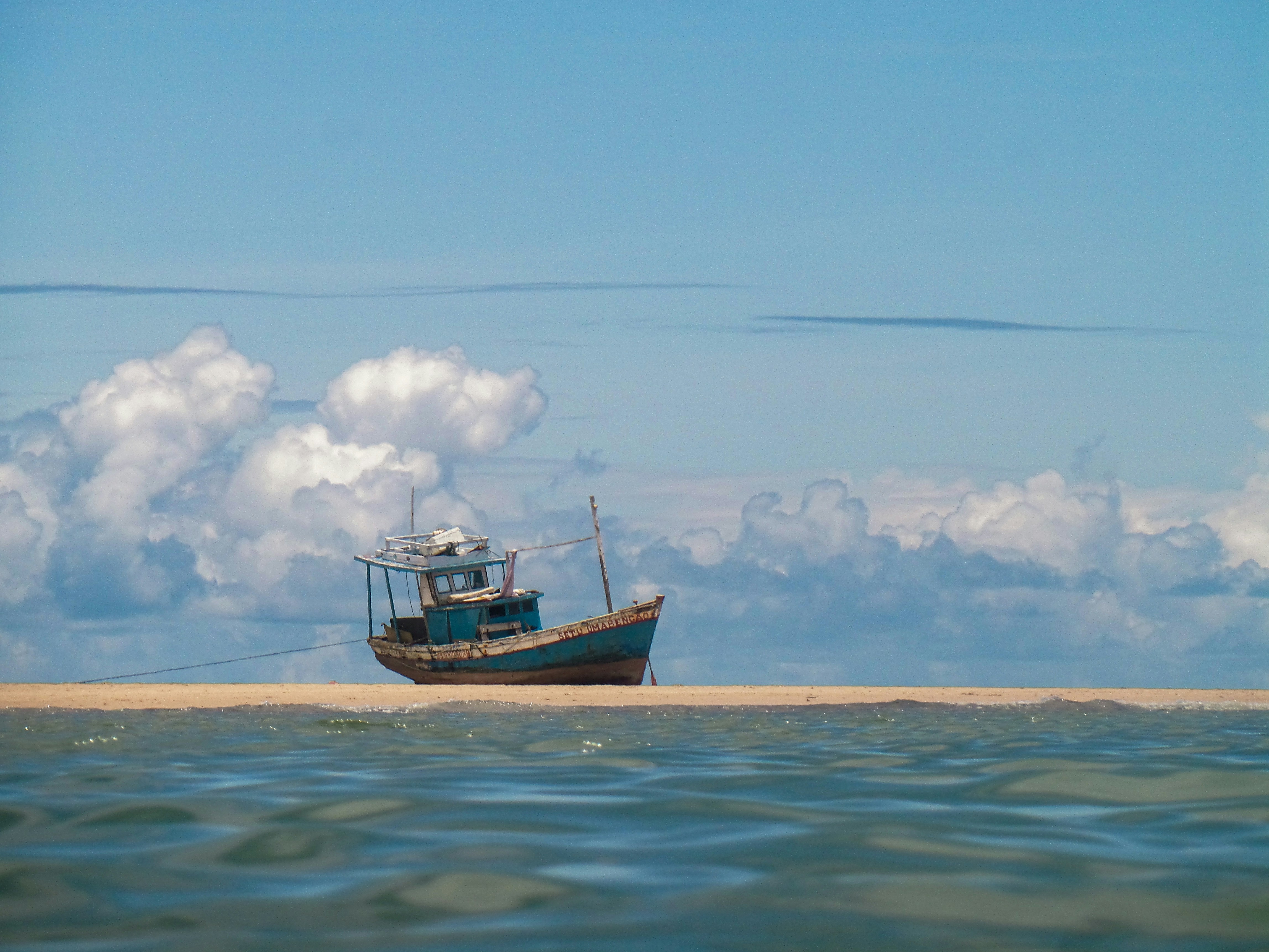 Abandoned fishing boat rests on a sandy shore under a vibrant blue sky with scattered clouds.