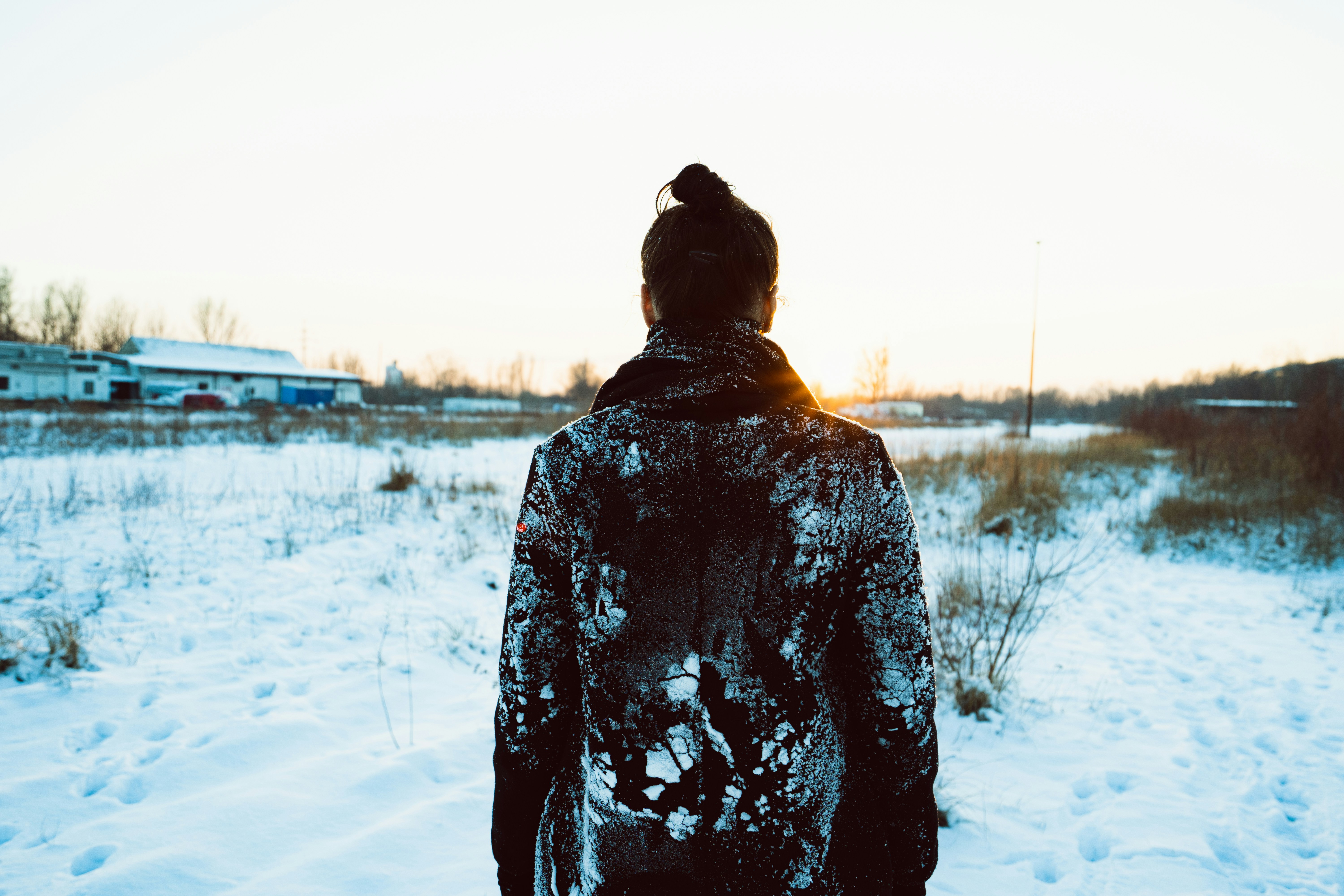 A figure stands in a snowy landscape, backlit by a soft sunset, wearing a coat with an artistic pattern. The serene atmosphere captures the essence of winter solitude.