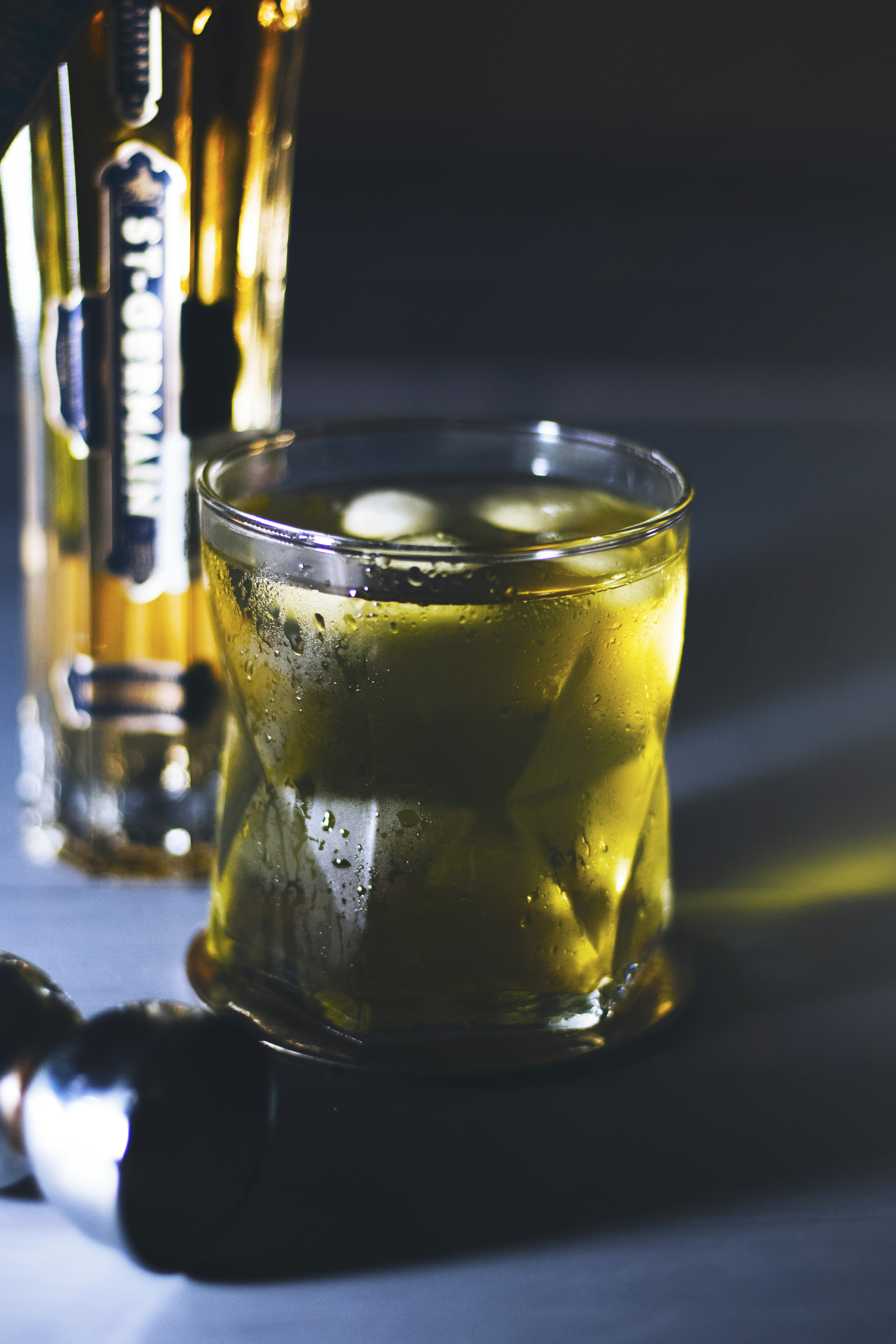 A glass of cocktail with ice, glistening droplets, and a bottle of St. Germain in the background, set against a dark surface.