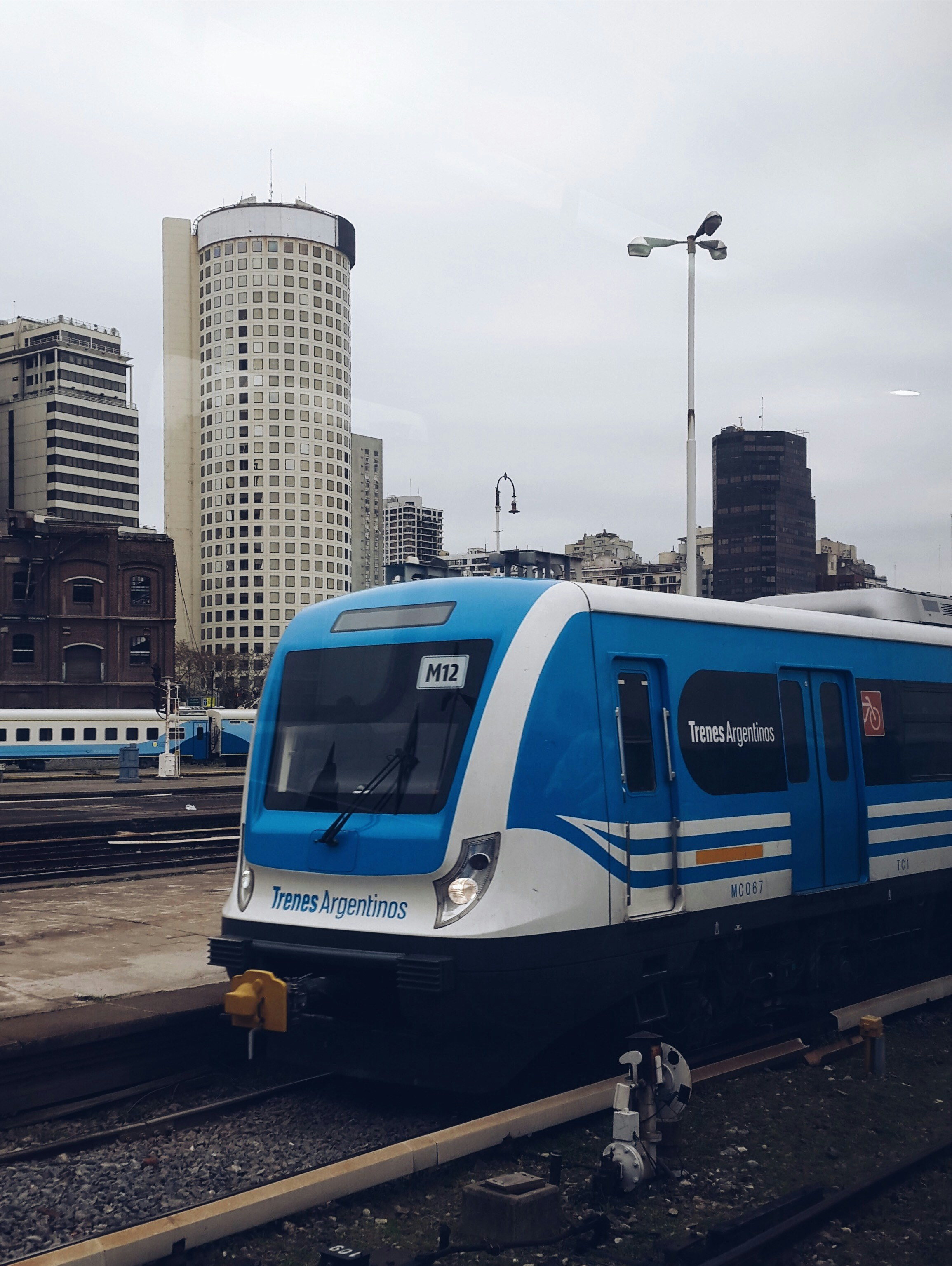 Blue Trenes Argentinos train glides along tracks with a city skyline in the background. The sleek front dominates the frame, conveying urban rail transit mood.