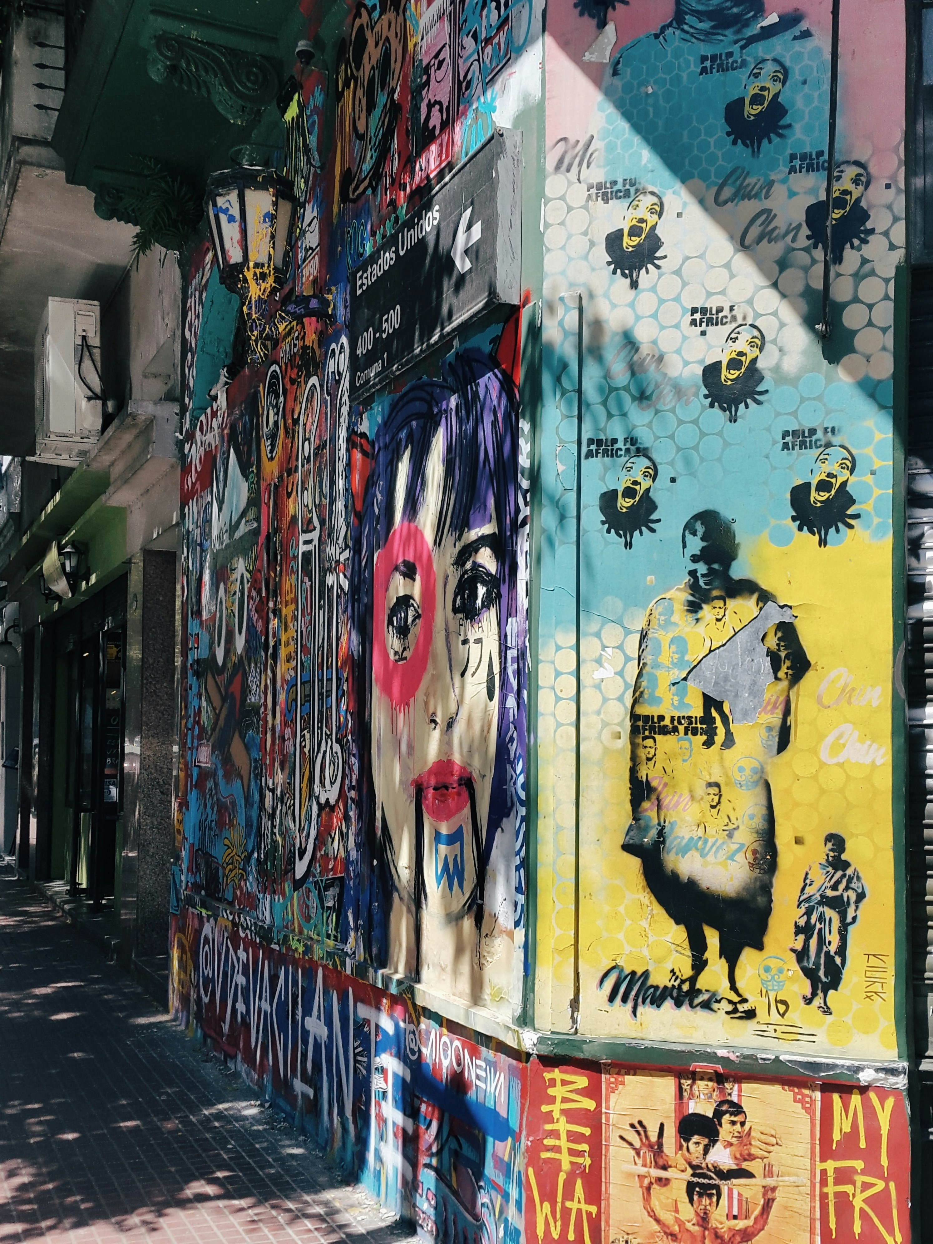 A sunlit alley wall crowded with vibrant street murals and posters, dominated by a large illustrated portrait with pink rings around the eyes.