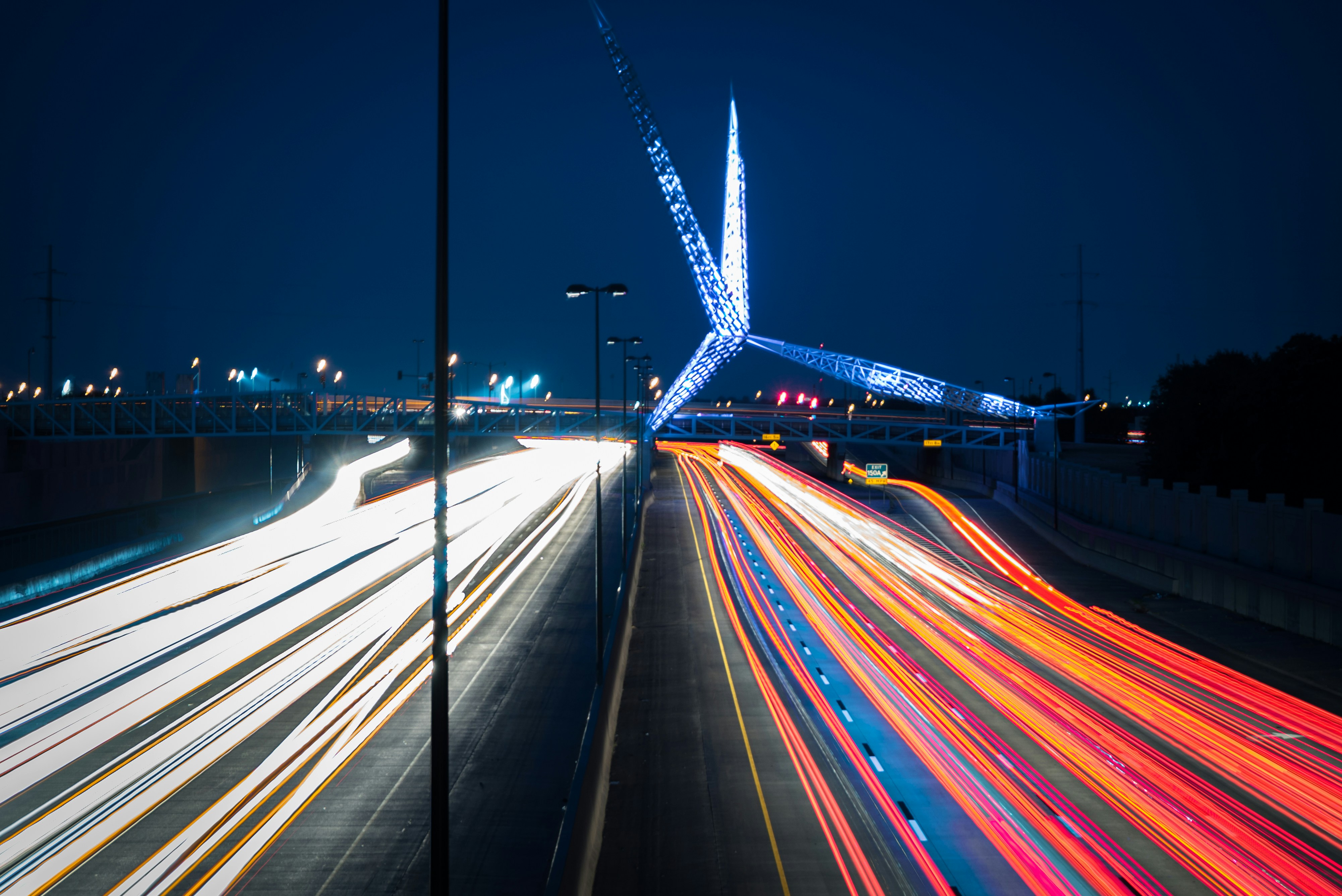 Long exposure of colorful car light trails on a highway beneath a modern bridge at night.